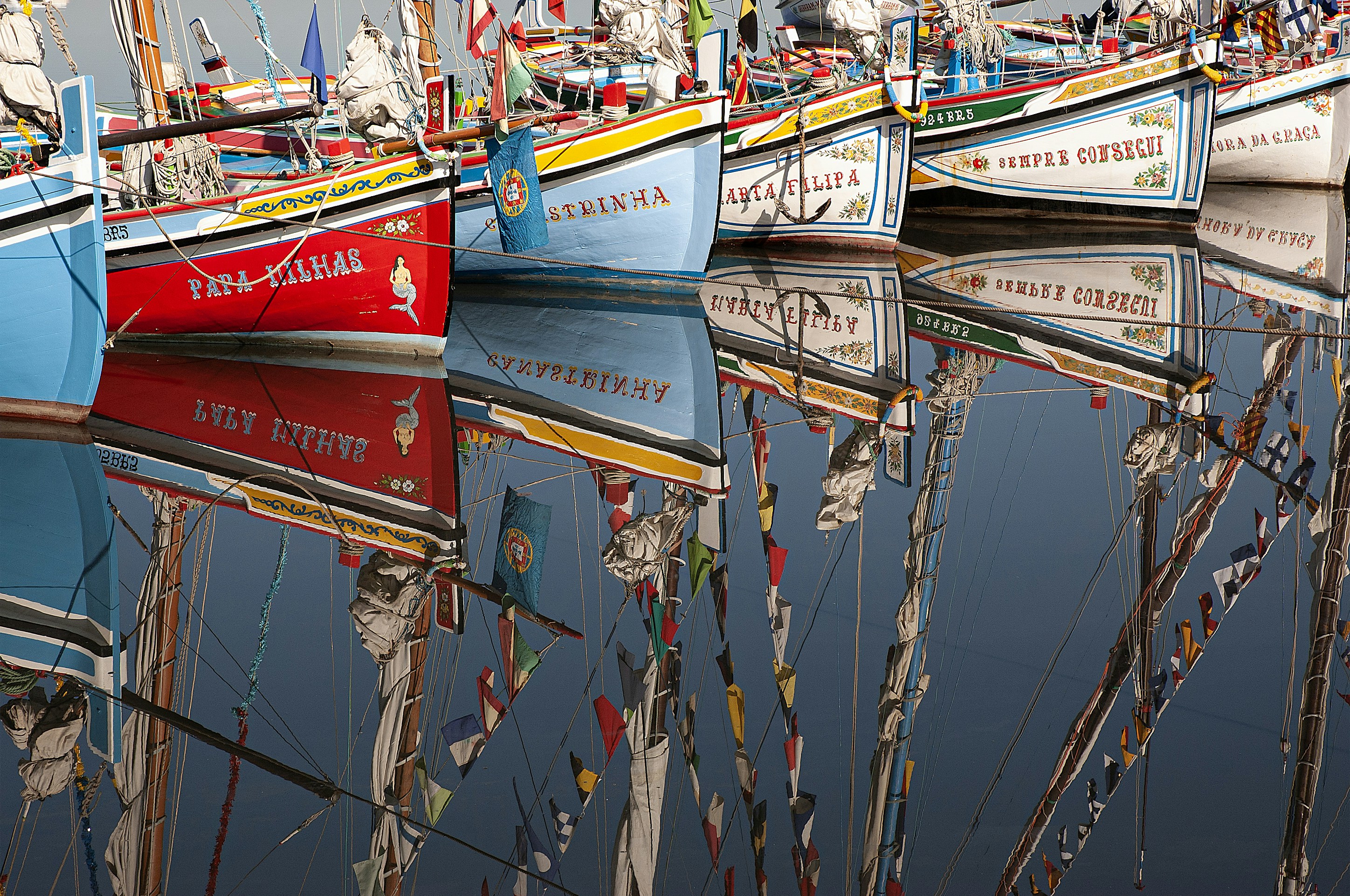 Colorful boats with banners line a harbor, their reflections perfectly mirrored in calm water, creating a geometric tapestry of lines and bright hues.