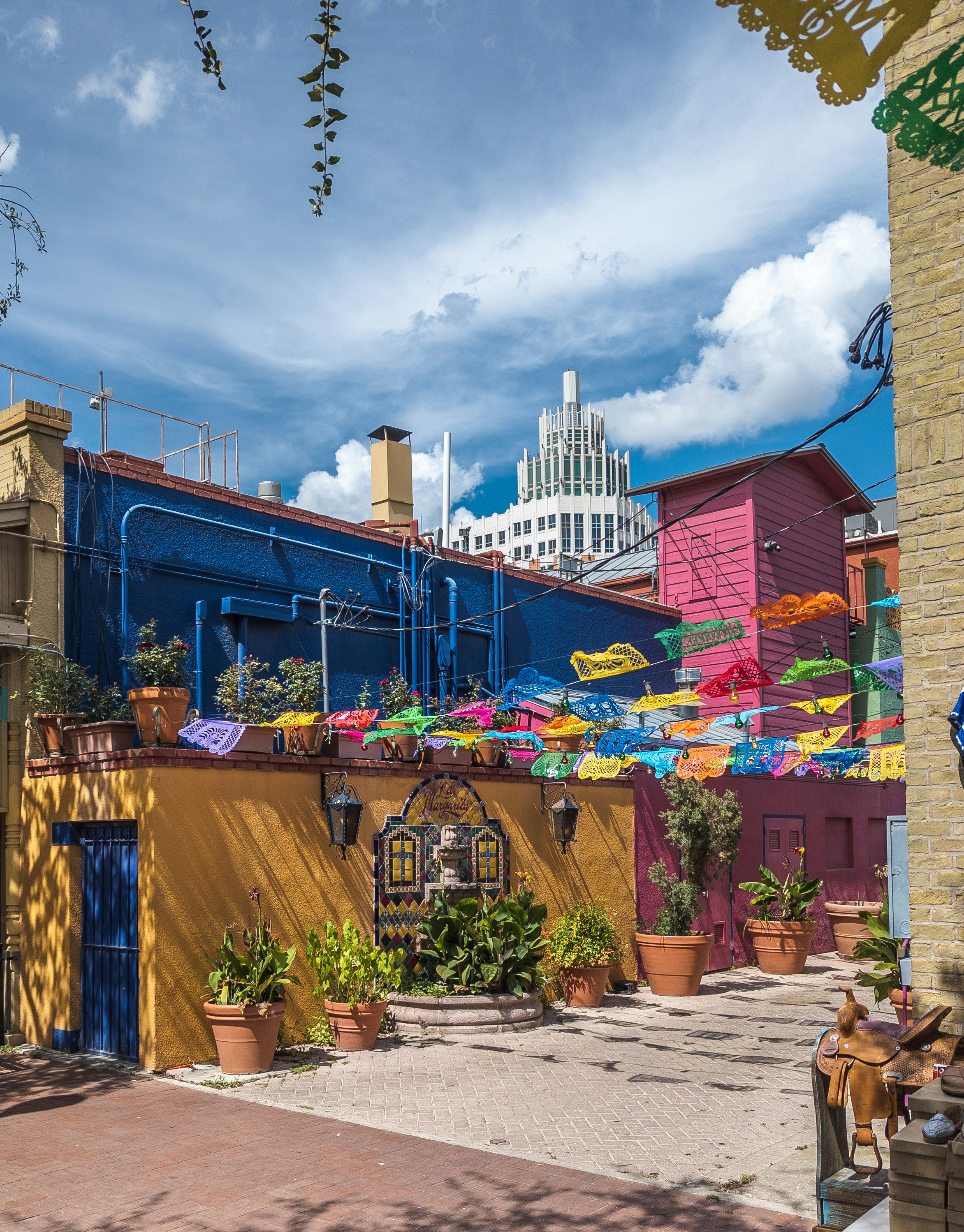 Vibrant courtyard lined with colorful walls and hanging banners, potted plants framing a sunlit paved space. A lively compilation of textures and colors evokes a festive neighborhood vibe.