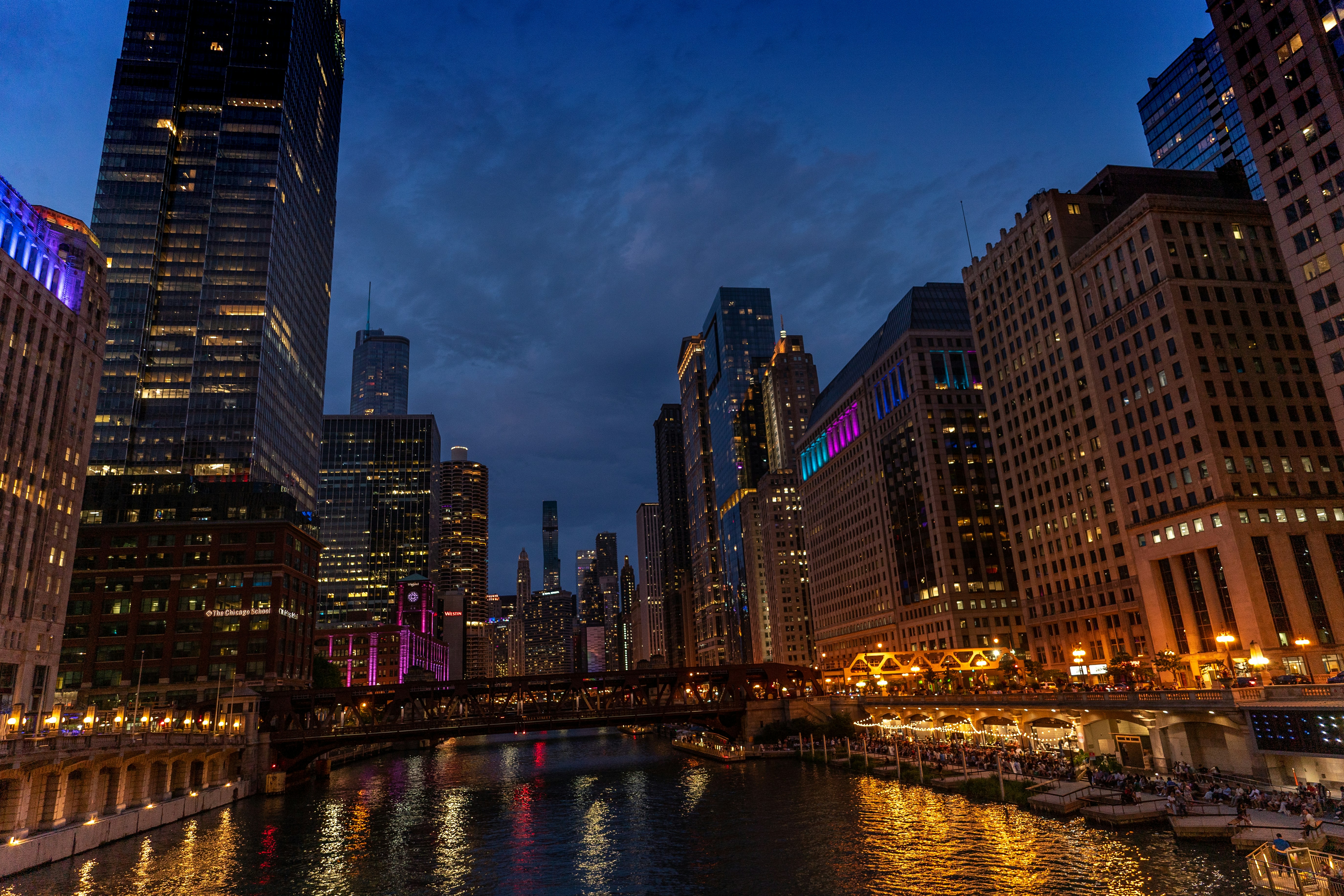 Nighttime cityscape with illuminated riverfront and bridge—tall buildings line the banks, reflections shimmer on the water, capturing a lively urban waterfront scene.