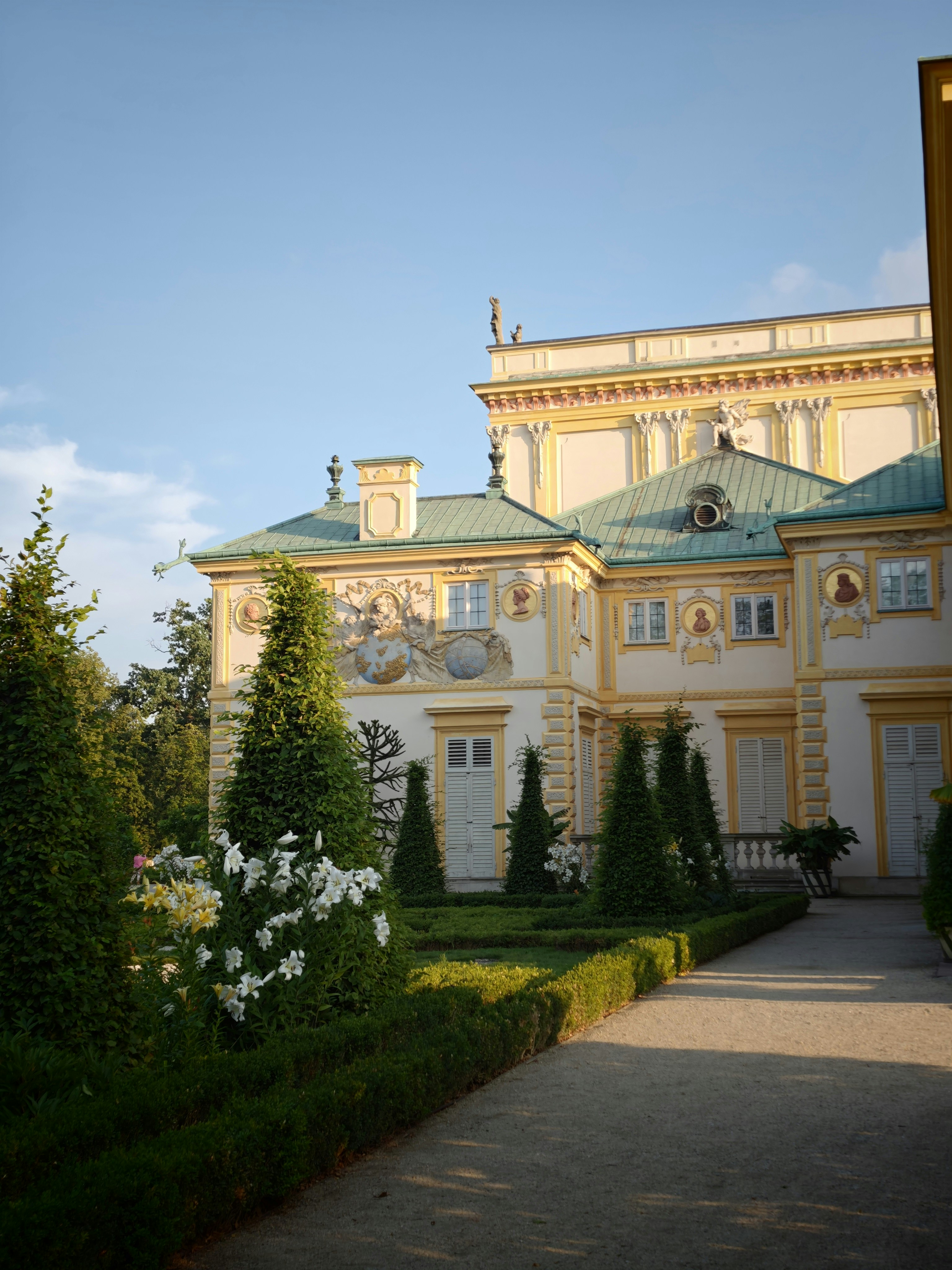 Elegant baroque palace façade bathed in afternoon light with neatly trimmed hedges and white flowers along a gravel path.