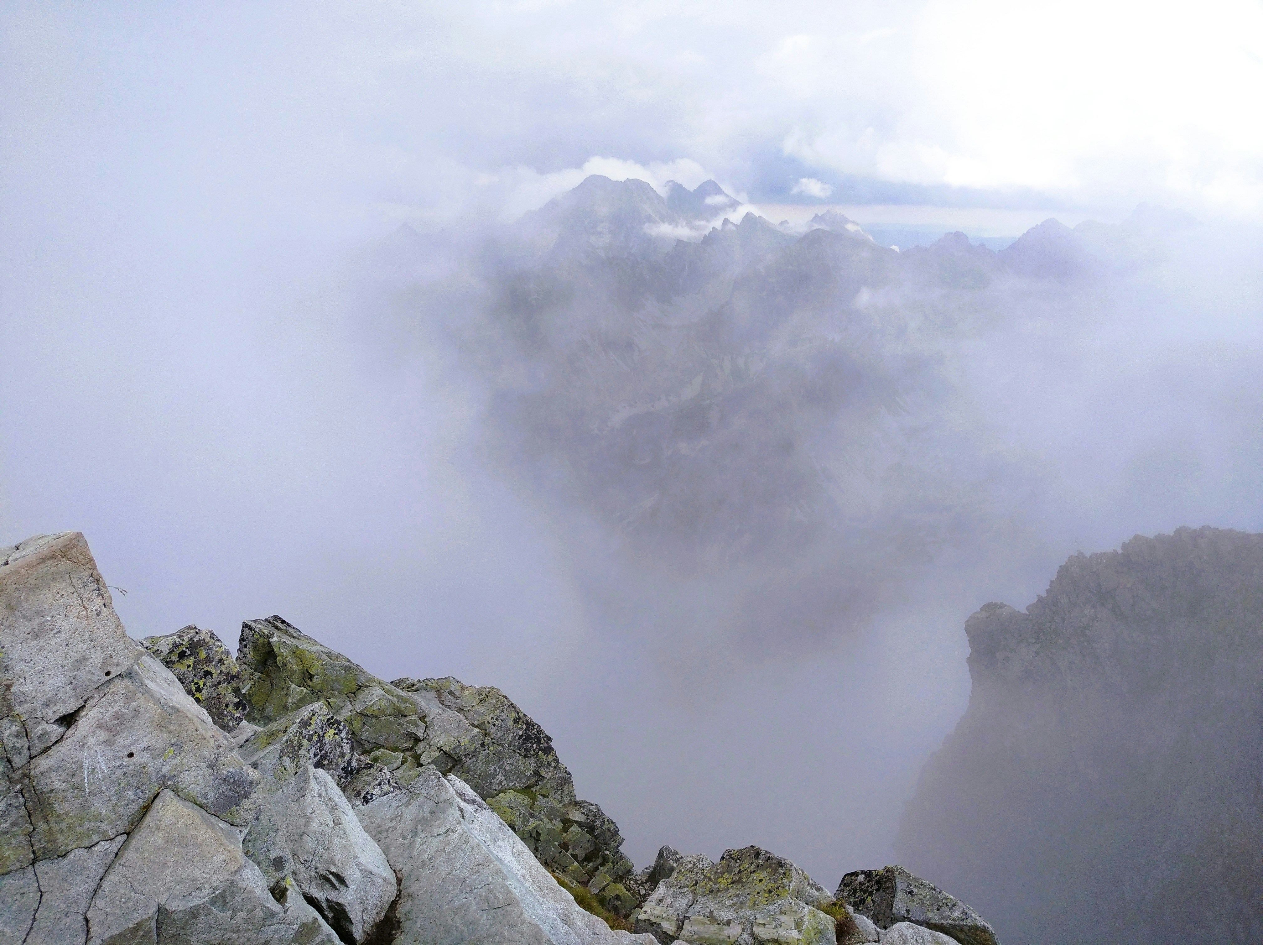 Cloud-enshrouded jagged ridge with distant, snow-topped peaks peeking through fog; a rugged alpine scene devoid of human presence. This is a landscape photograph.