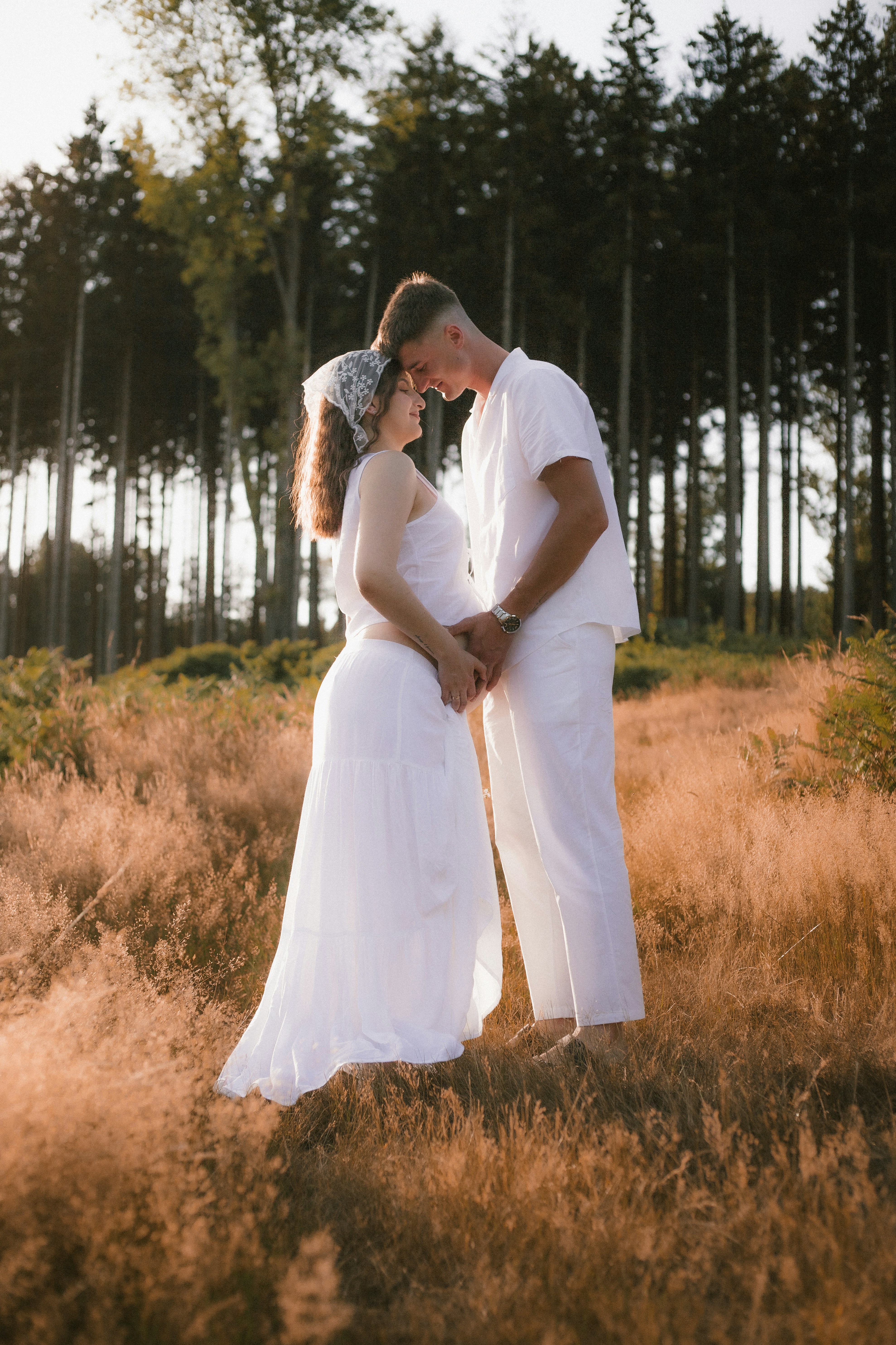White-dressed couple in sunlit meadow share a quiet moment by the pines