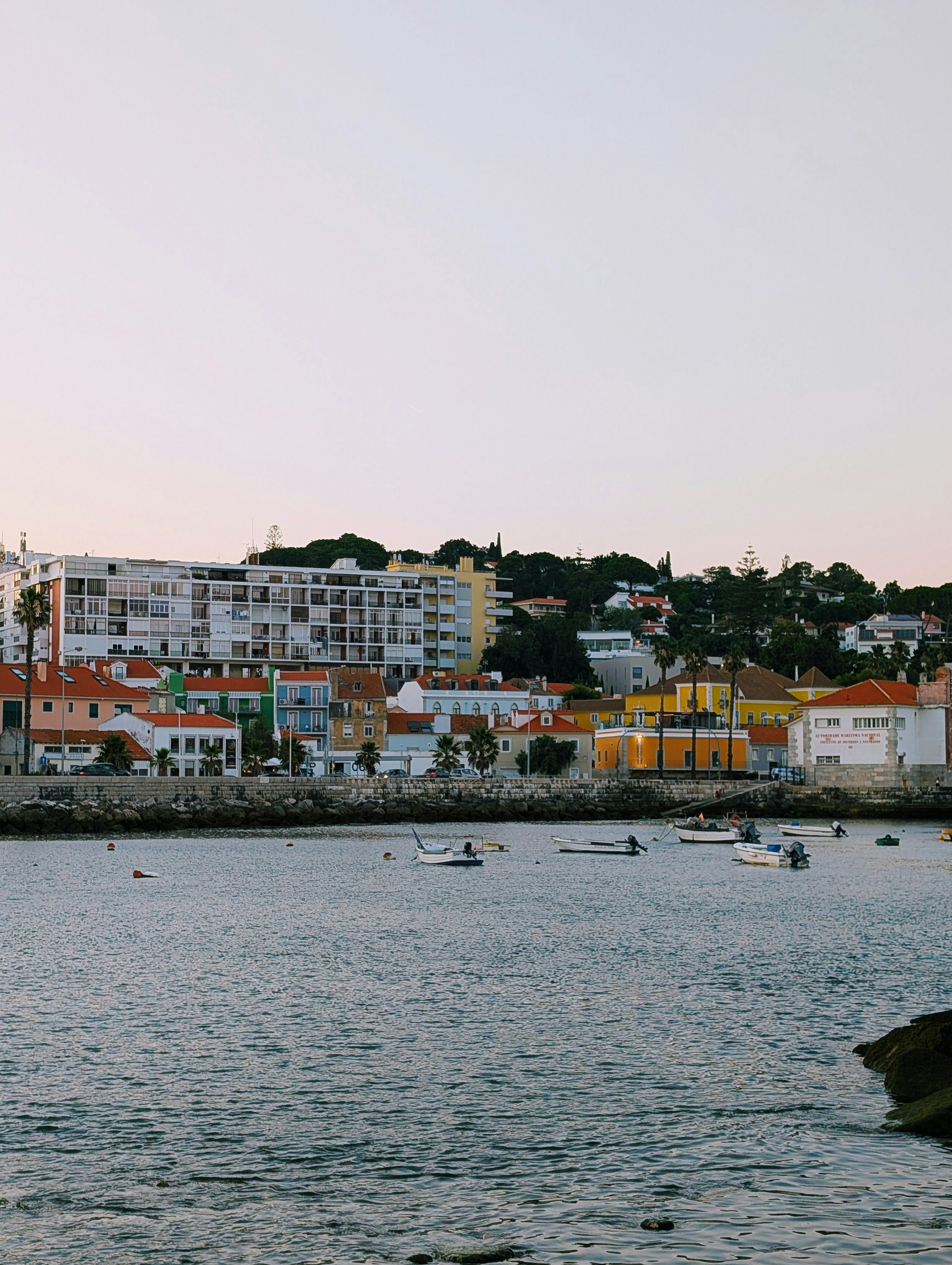 Colorful houses line the waterfront with boats dotting the calm harbor; the scene shows a quiet coastal town with a soft evening sky.