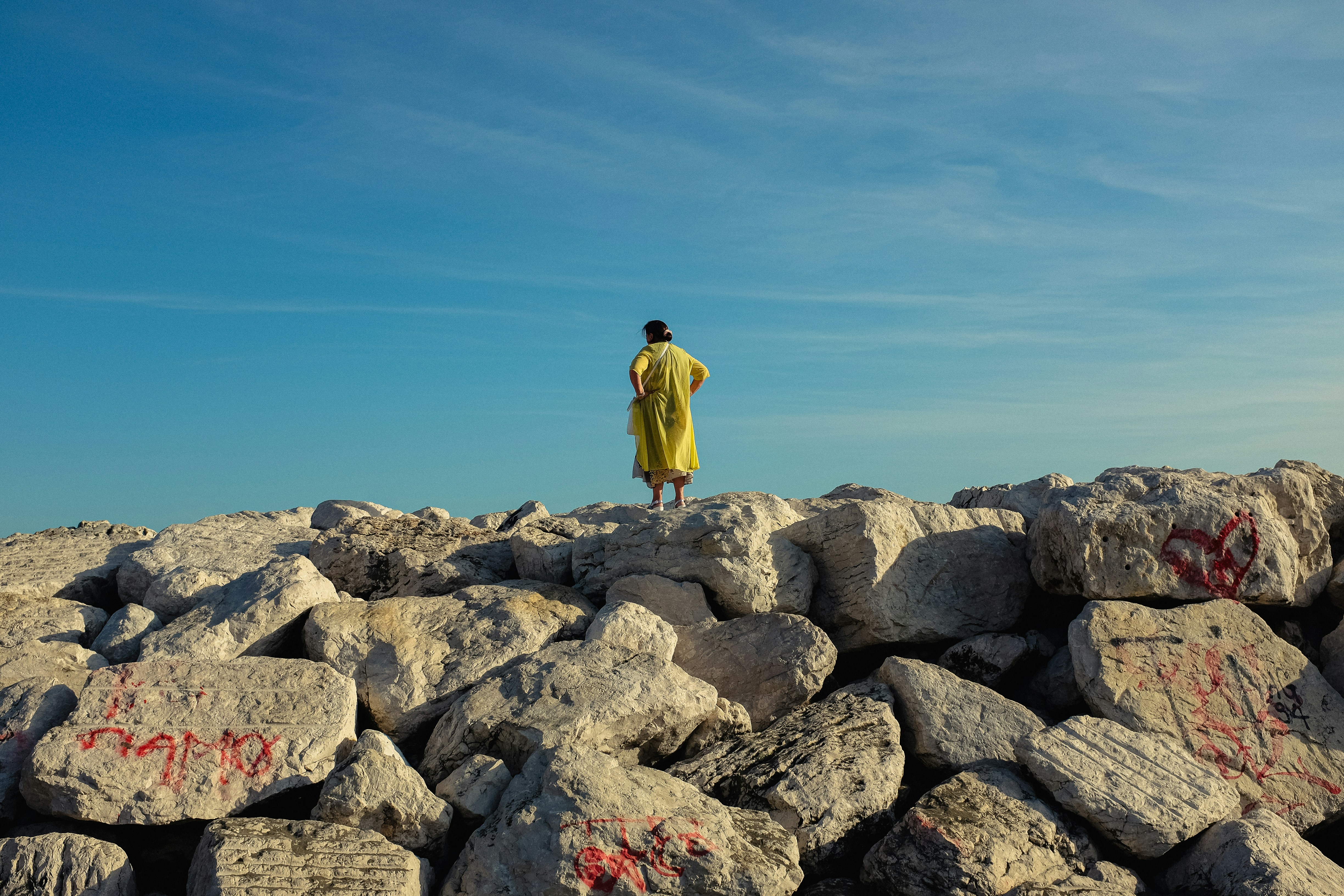 Yellow-clad figure surveying a rocky breakwater under a vast blue sky