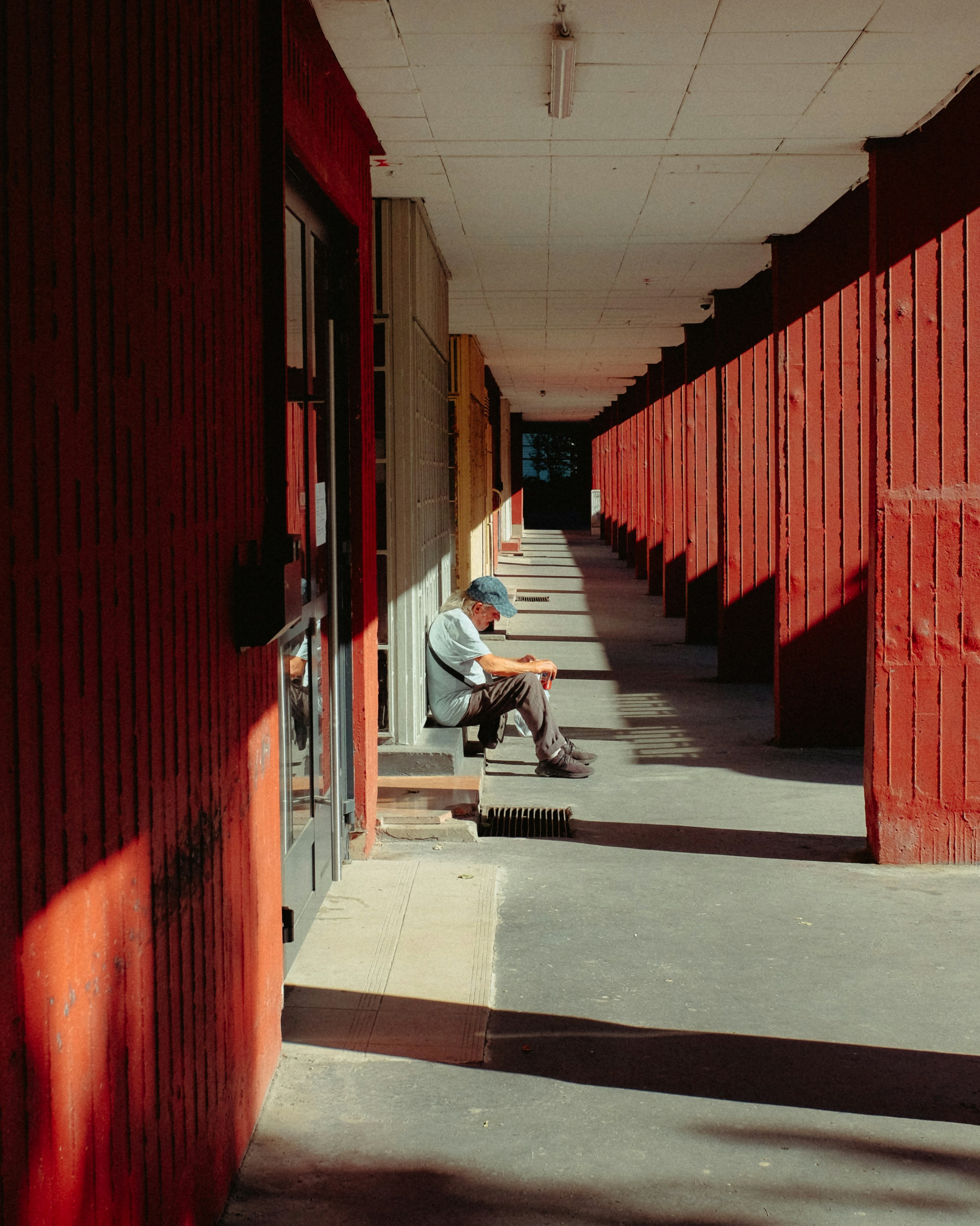 A man sits and reads under a shaded arcade of red columns, casting long geometric shadows along a concrete walkway.