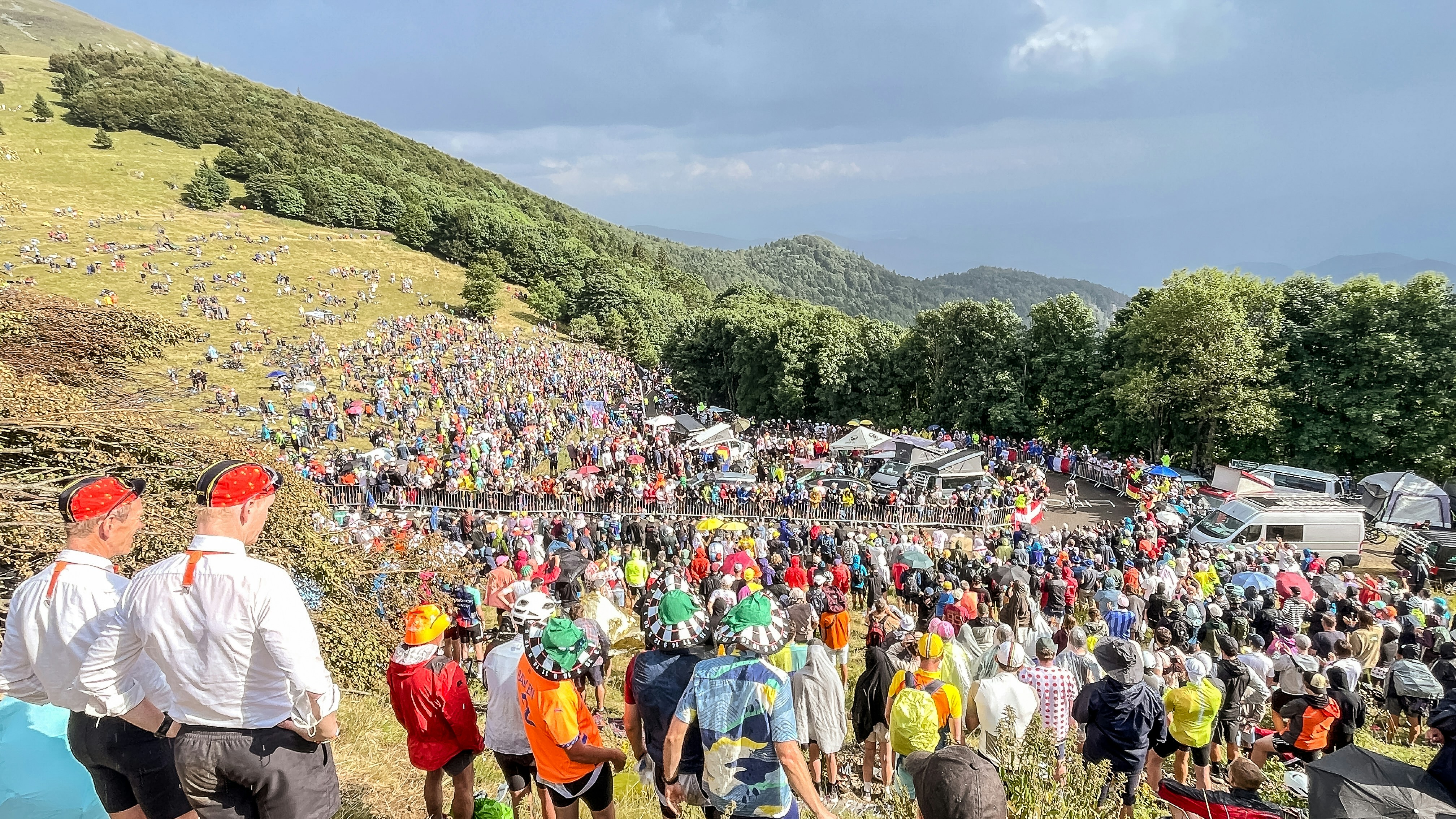Vibrant crowd gathers on a hillside at a communal event with tents and vehicles at the base; a sea of people in colorful outfits creates a lively scene.
