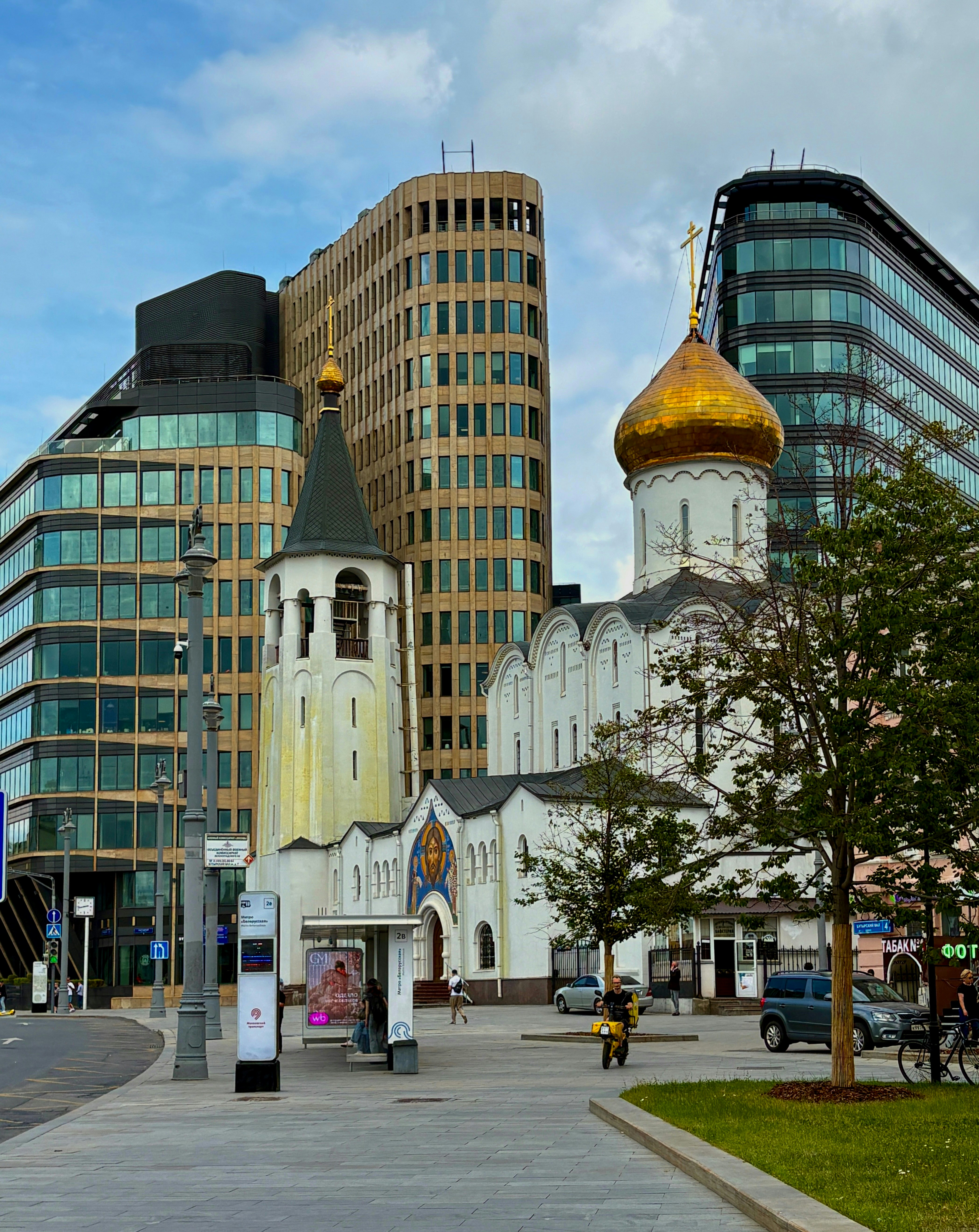 Dome-roofed church sits at the edge of a modern glass-fronted boulevard, blending historic architecture with contemporary buildings in a bright urban square.