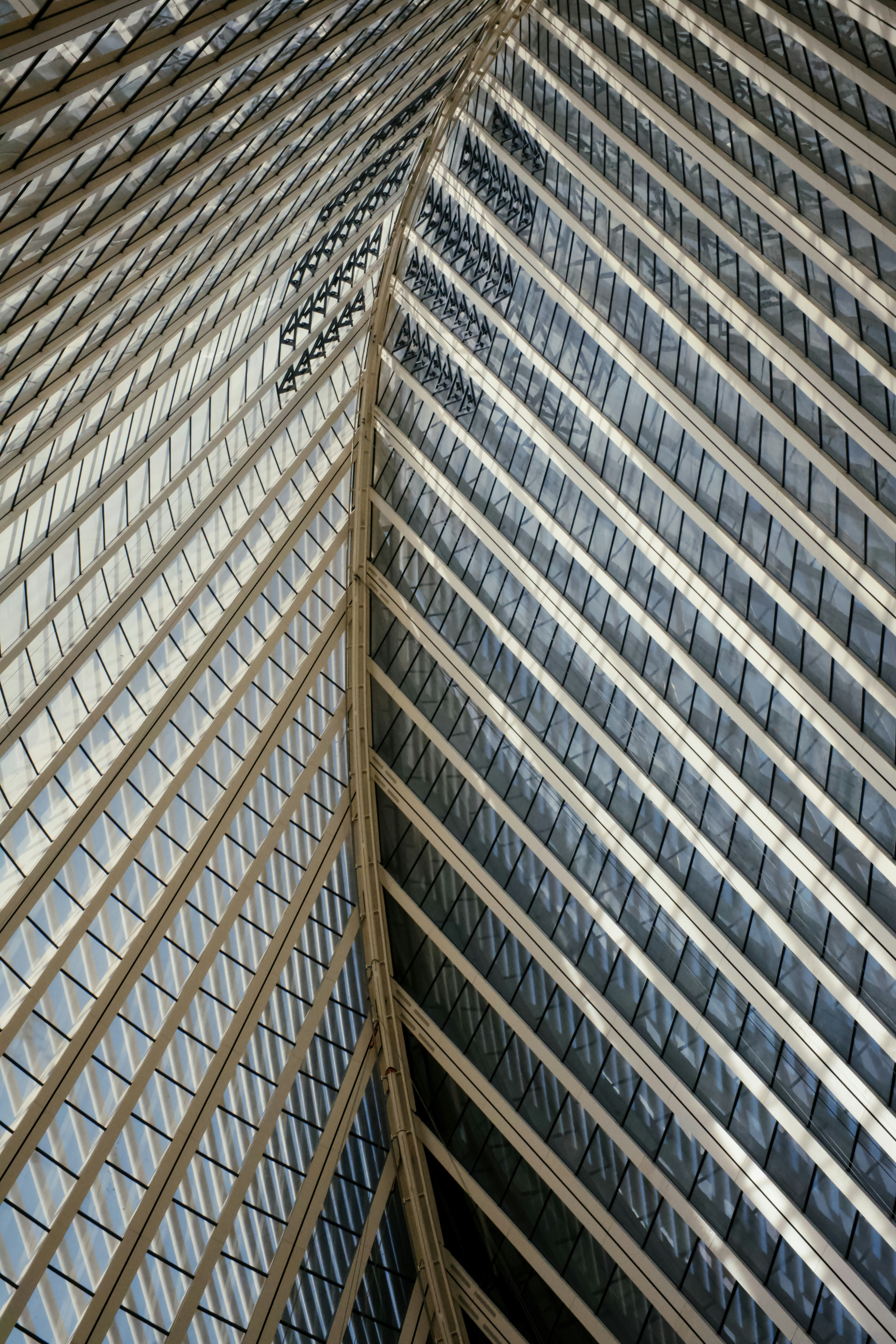 Converging diagonal ribs of a modern glass-and-metal structure create a dynamic, geometric pattern with cool blue reflections and warm beige supports.