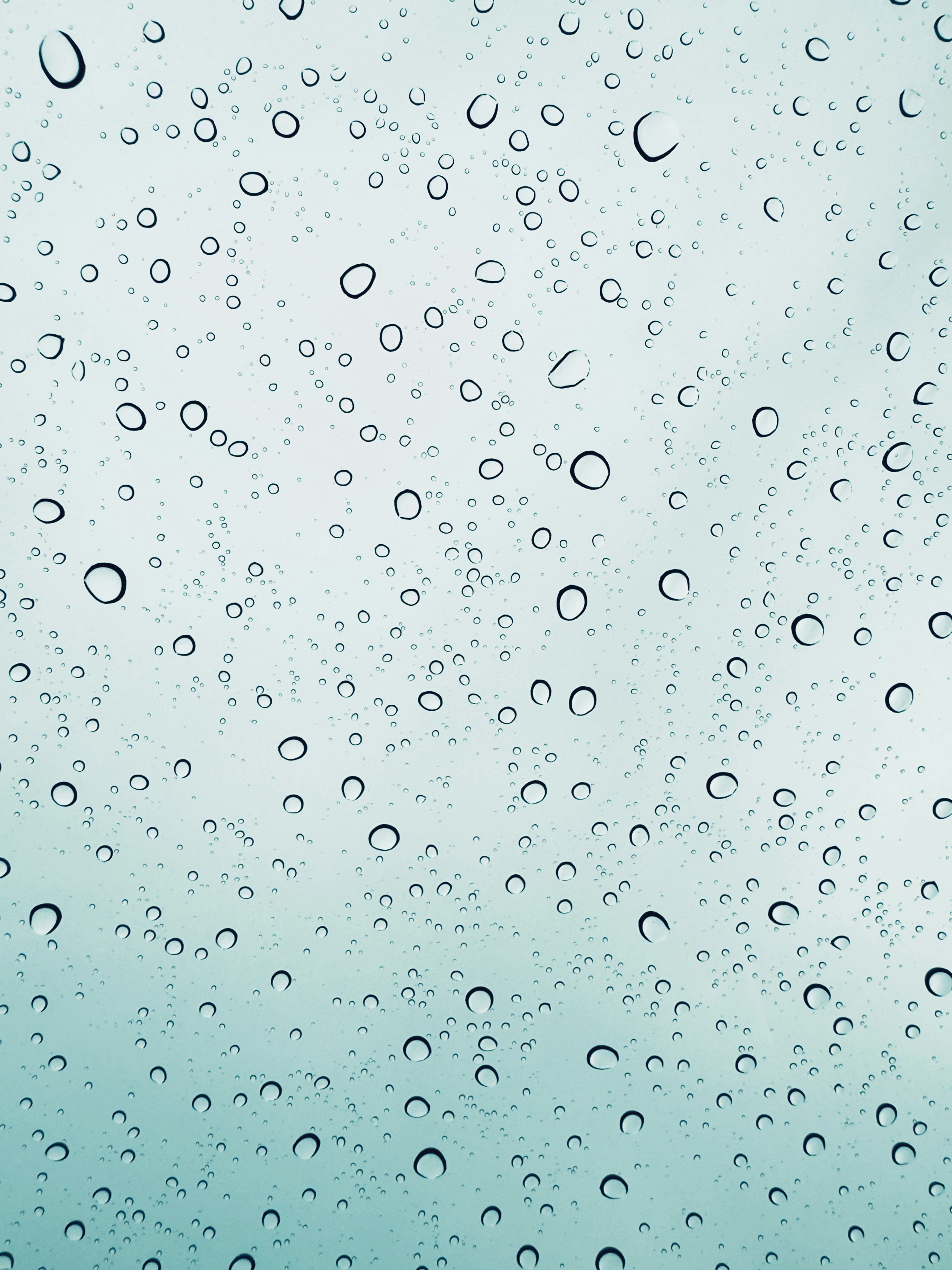 Close-up of numerous water droplets clinging to a window pane, creating a soft gradient background of cool blue-green tones.
