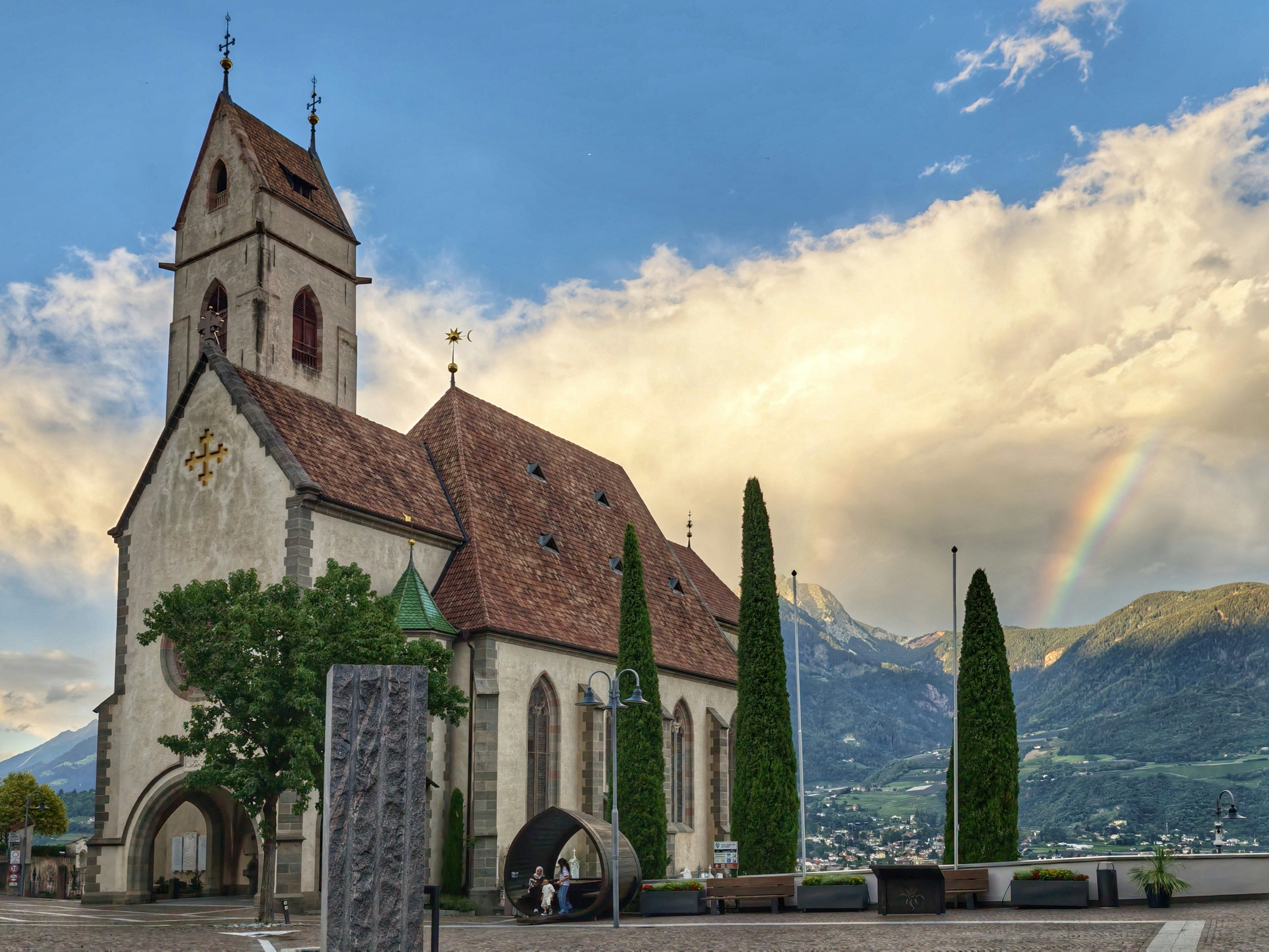 Alpine church with a steep roof sits beside a calm lake, framed by tall cypress-like trees and distant mountains under a partly cloudy sky with a rainbow.