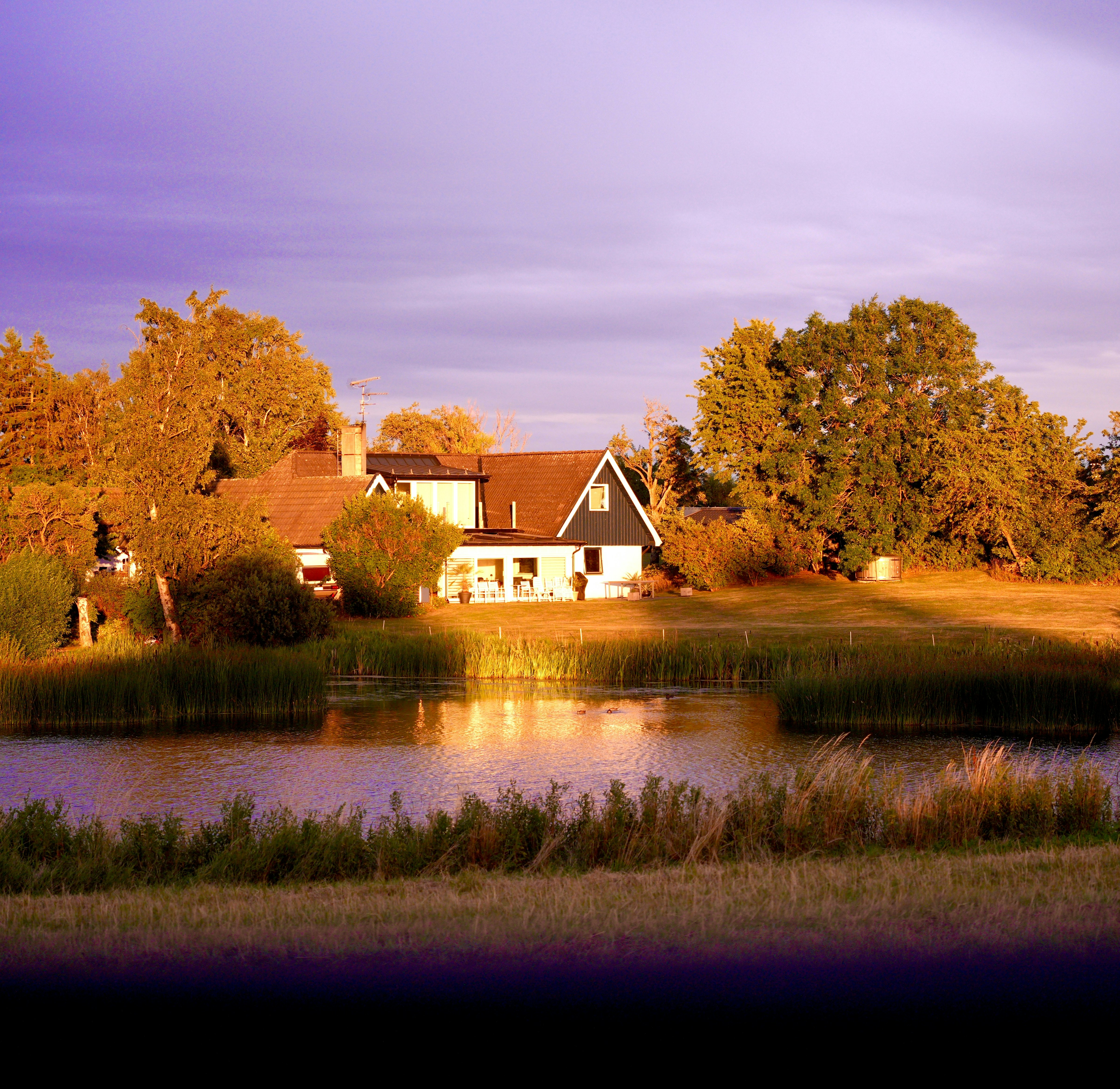 Warm-toned riverside cottage bathed in golden light with trees framing a calm waterway and a grassy foreground.
