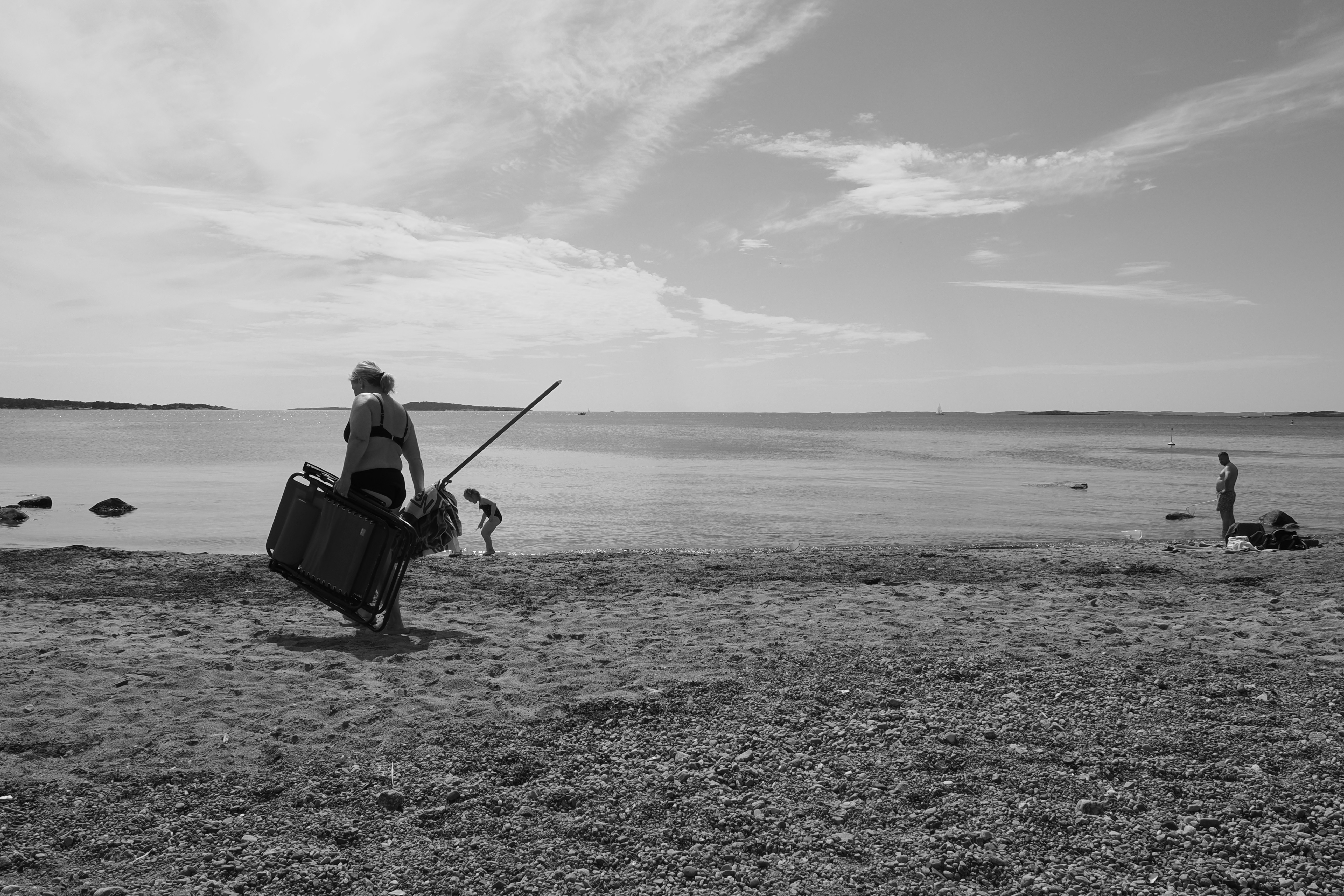 People on a pebbled beach with a person carrying a large suitcase toward the water; a small boat and a distant figure add depth to a calm seascape.