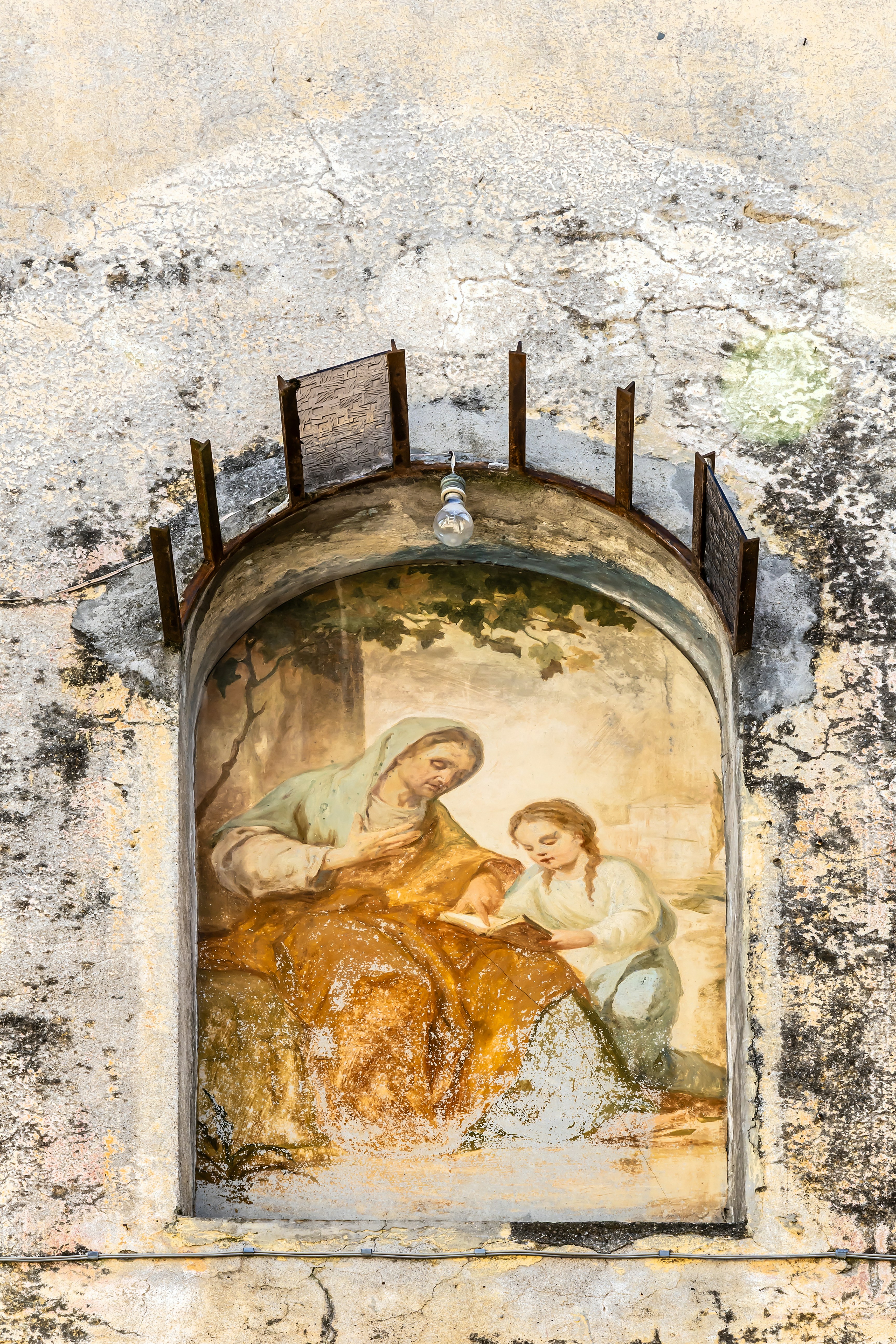 Painted religious scene framed by a rounded alcove, set in a weathered exterior wall; a single bulb hangs above, casting soft light on the artwork.