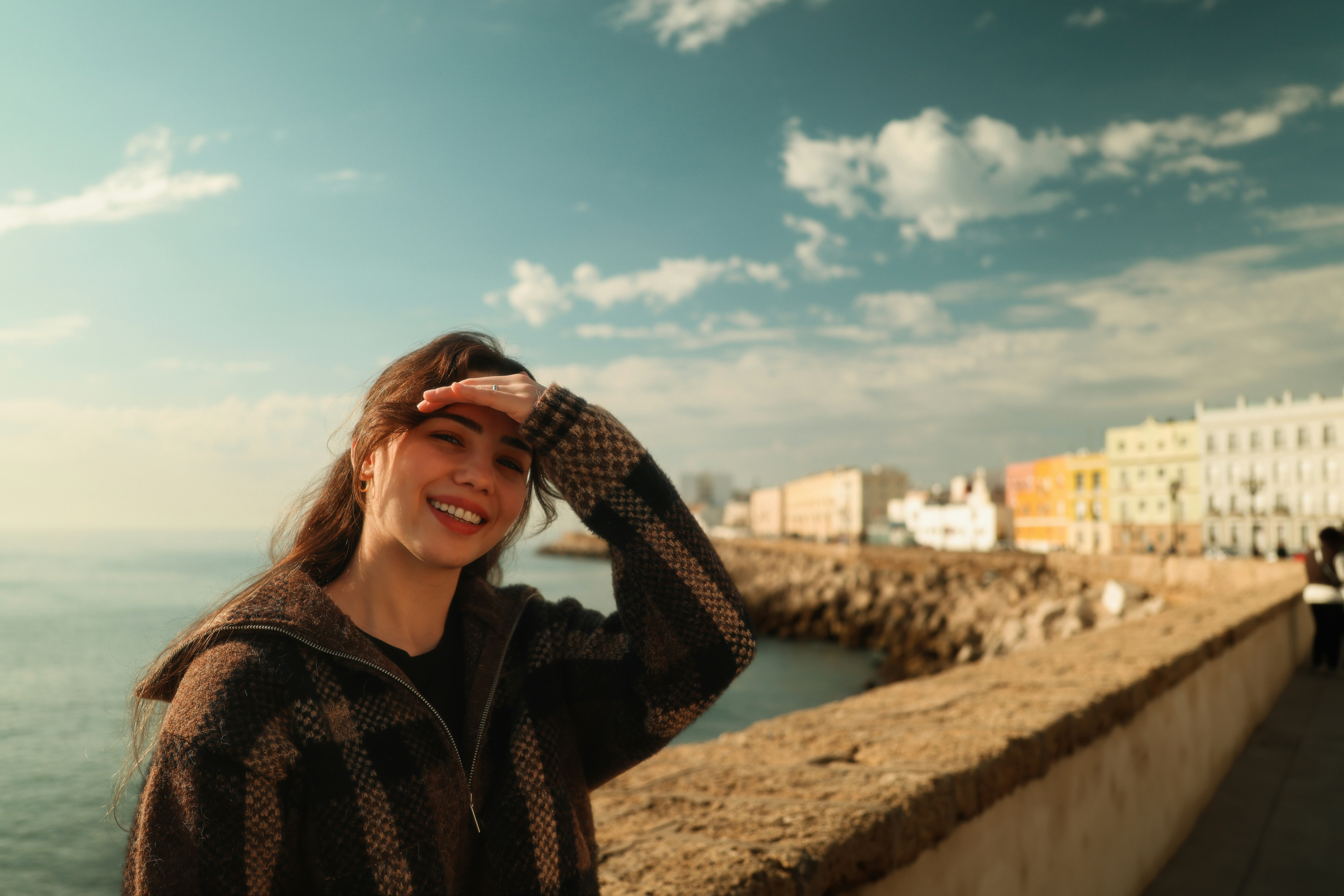 Smiling woman shielding her eyes with a hand along a sunlit seaside promenade; colorful buildings line the harbor in the background.