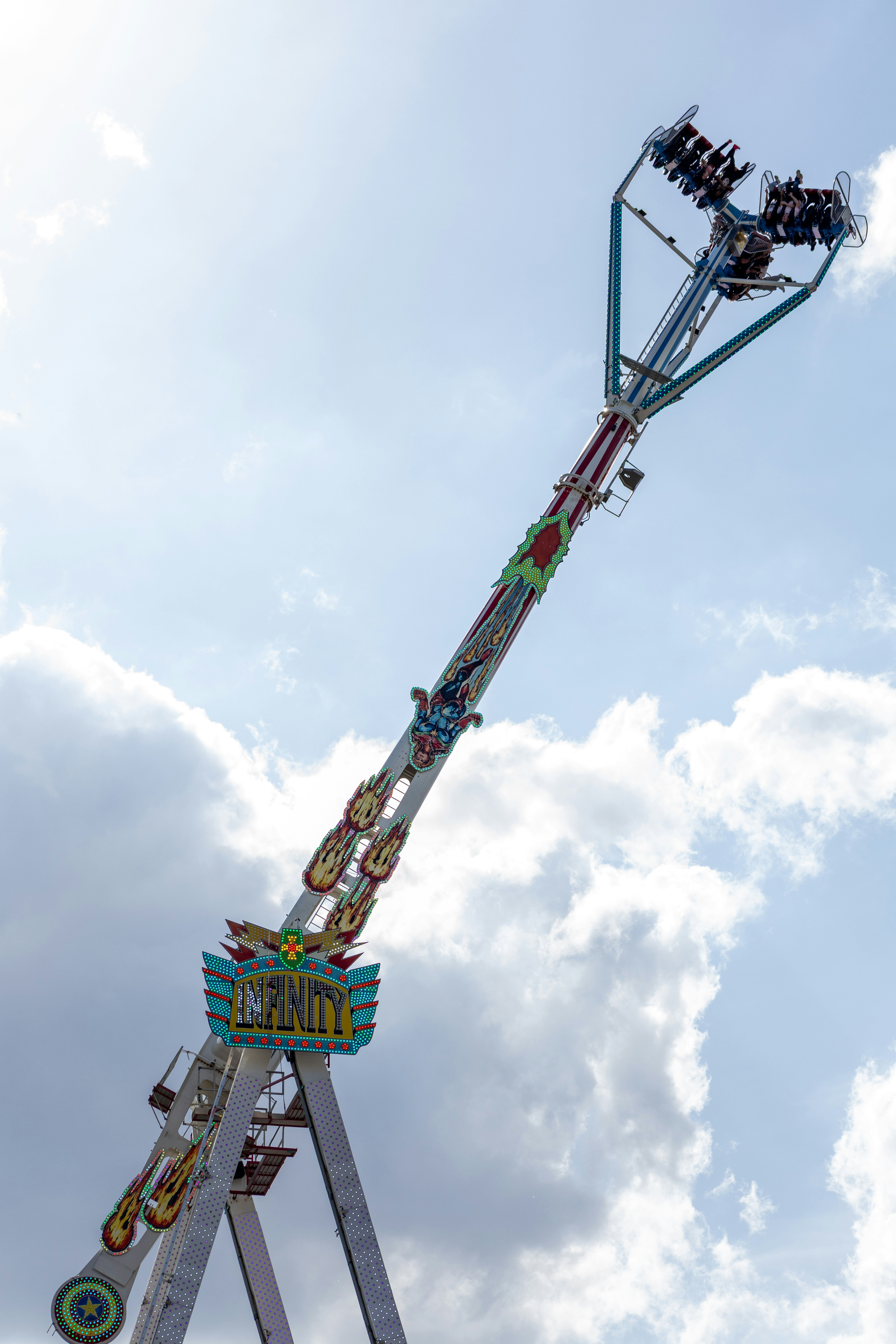 A towering amusement ride ascends into a bright sky, with riders silhouetted at the top as the arm stretches upward; a colorful sign and ornate artwork run along the supporting structure. This is a high-energy, mobile amusement environment.