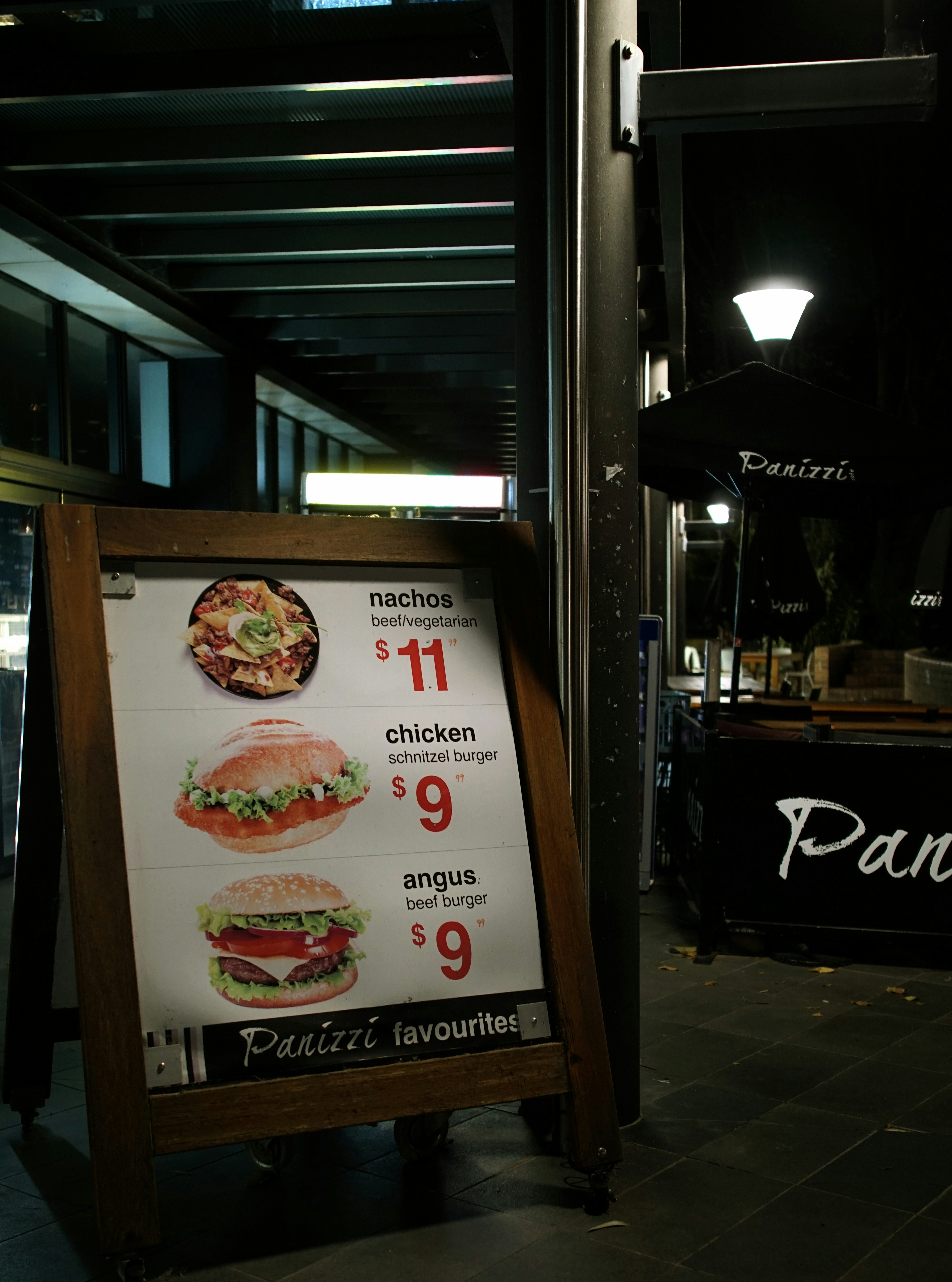 A lit sandwich-board menu stands on a dim sidewalk near a restaurant exterior, showing images and prices for nachos, chicken, and angus burgers beside an illuminated street scene.