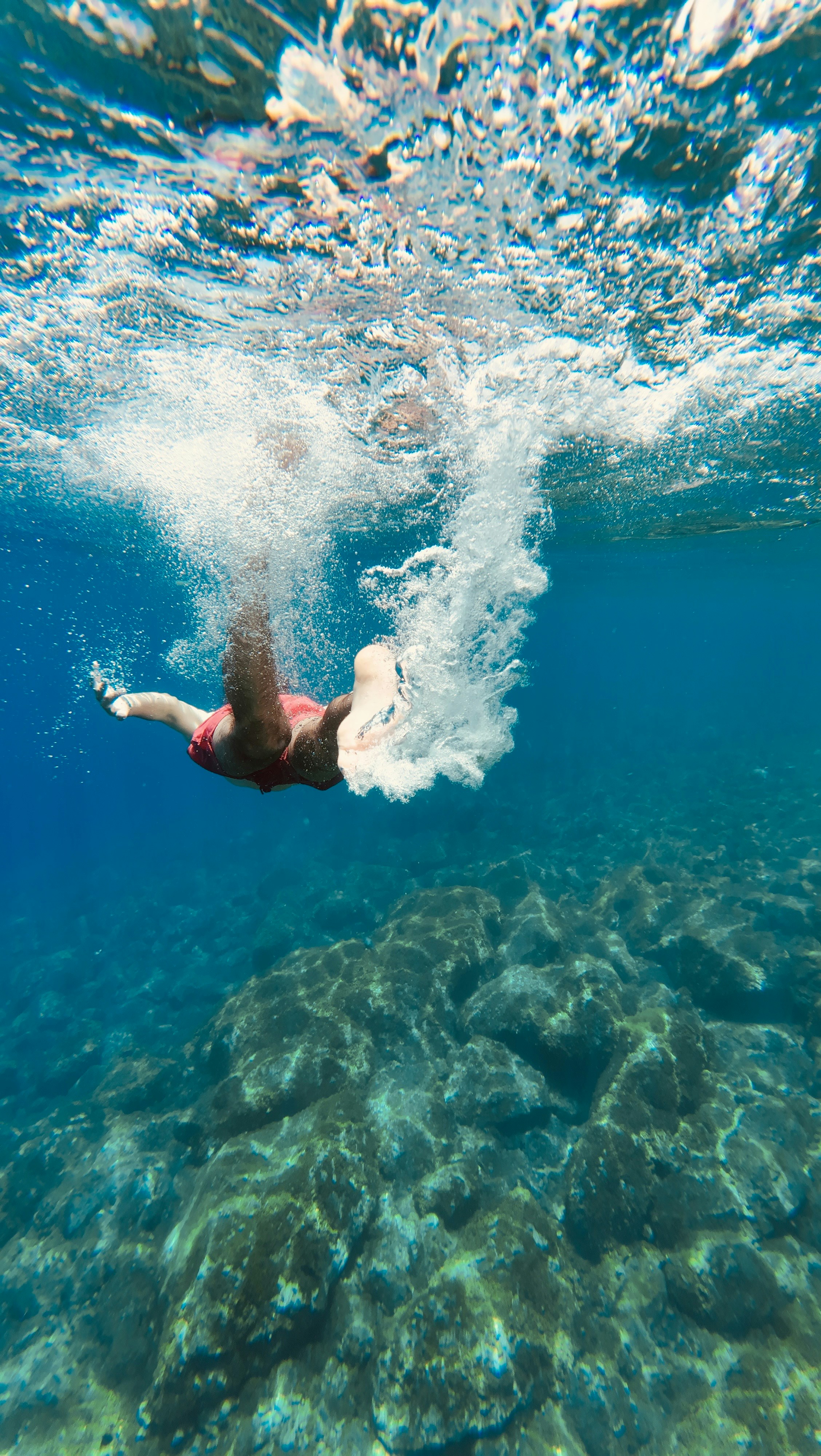 A swimmer dives beneath the surface, red swimsuit contrasting with the blue-green ocean and rocky seabed visible through crystal water.