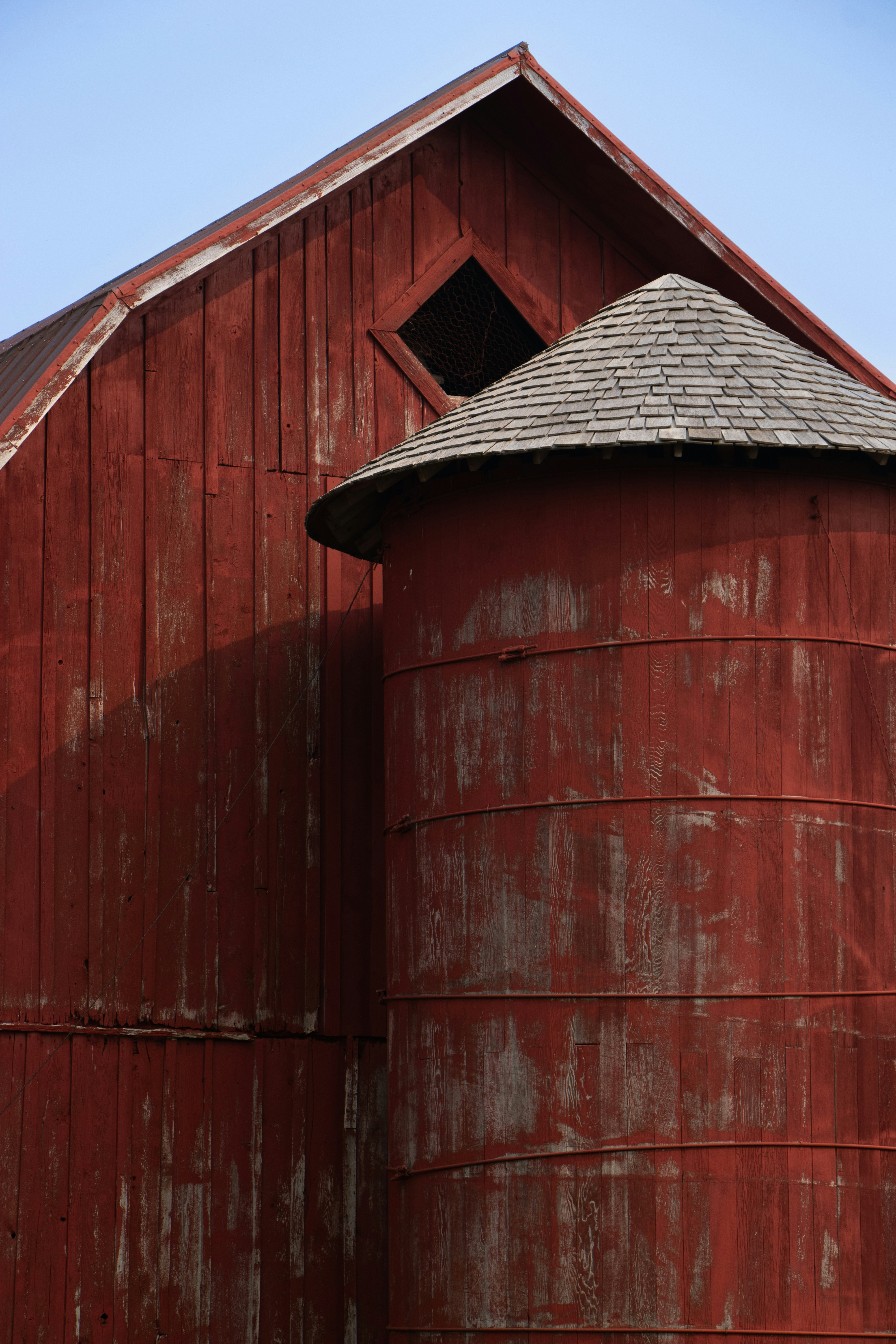 Weathered red barn with a curved silo dominates the frame, roof peaks cut a bold geometric line against a clear sky. A textured, aging surface emphasizes rural architecture. Painted wood and metal surfaces mark years of exposure.