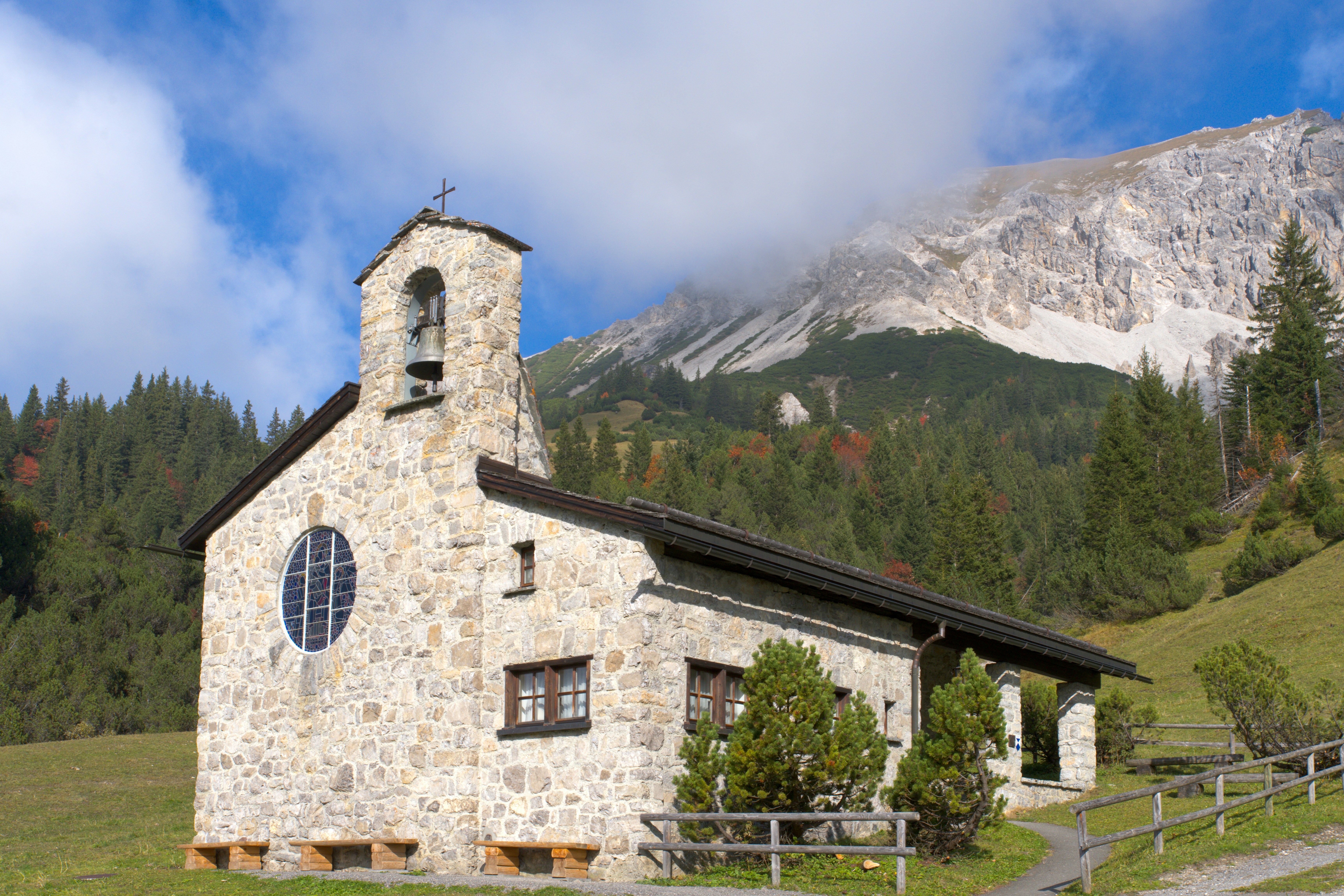 A small stone chapel set in a sunlit alpine meadow with a wooden fence and mountains in the background. The scene emphasizes rustic architecture and serene landscape.