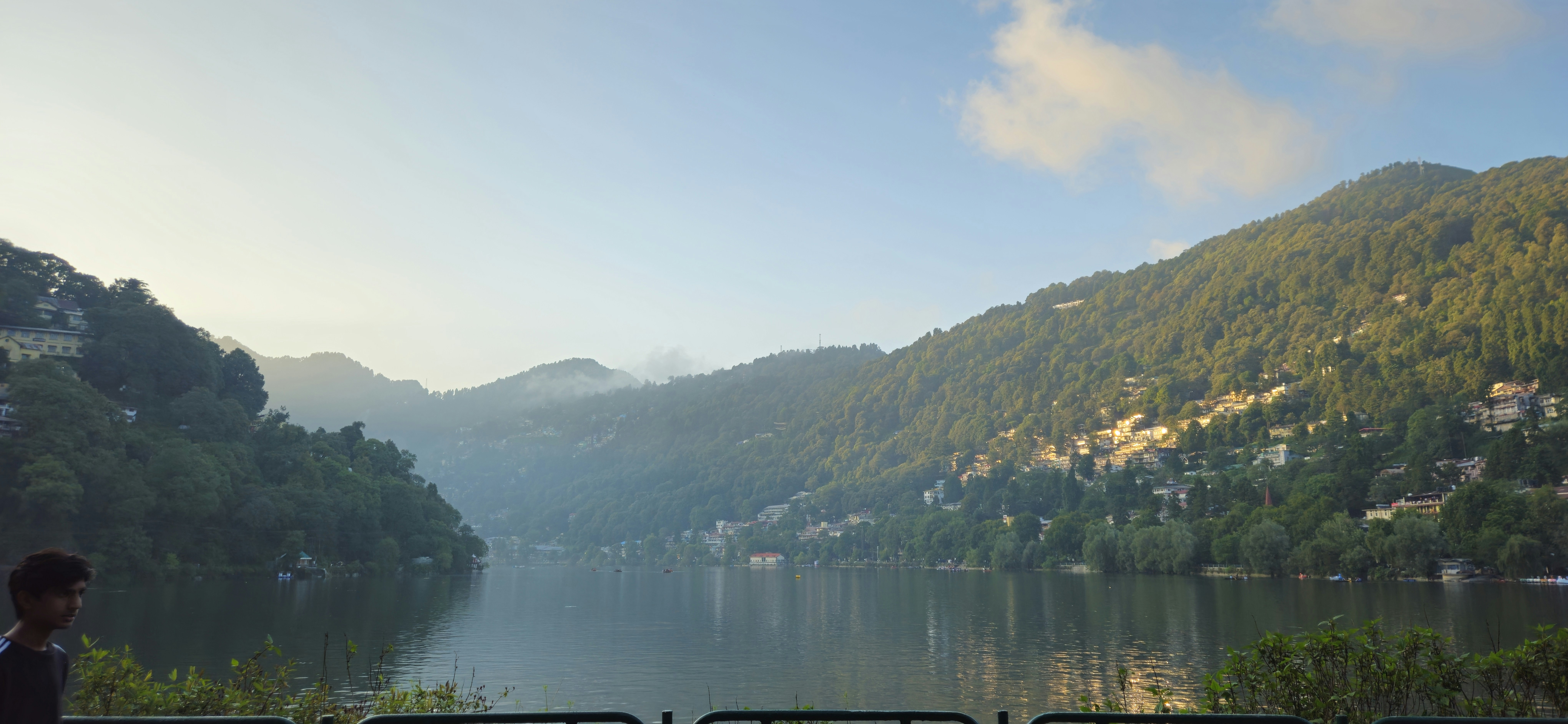 Tranquil lake flanked by tree-covered mountains under a clear sky; distant town lights begin to glow along the shore.