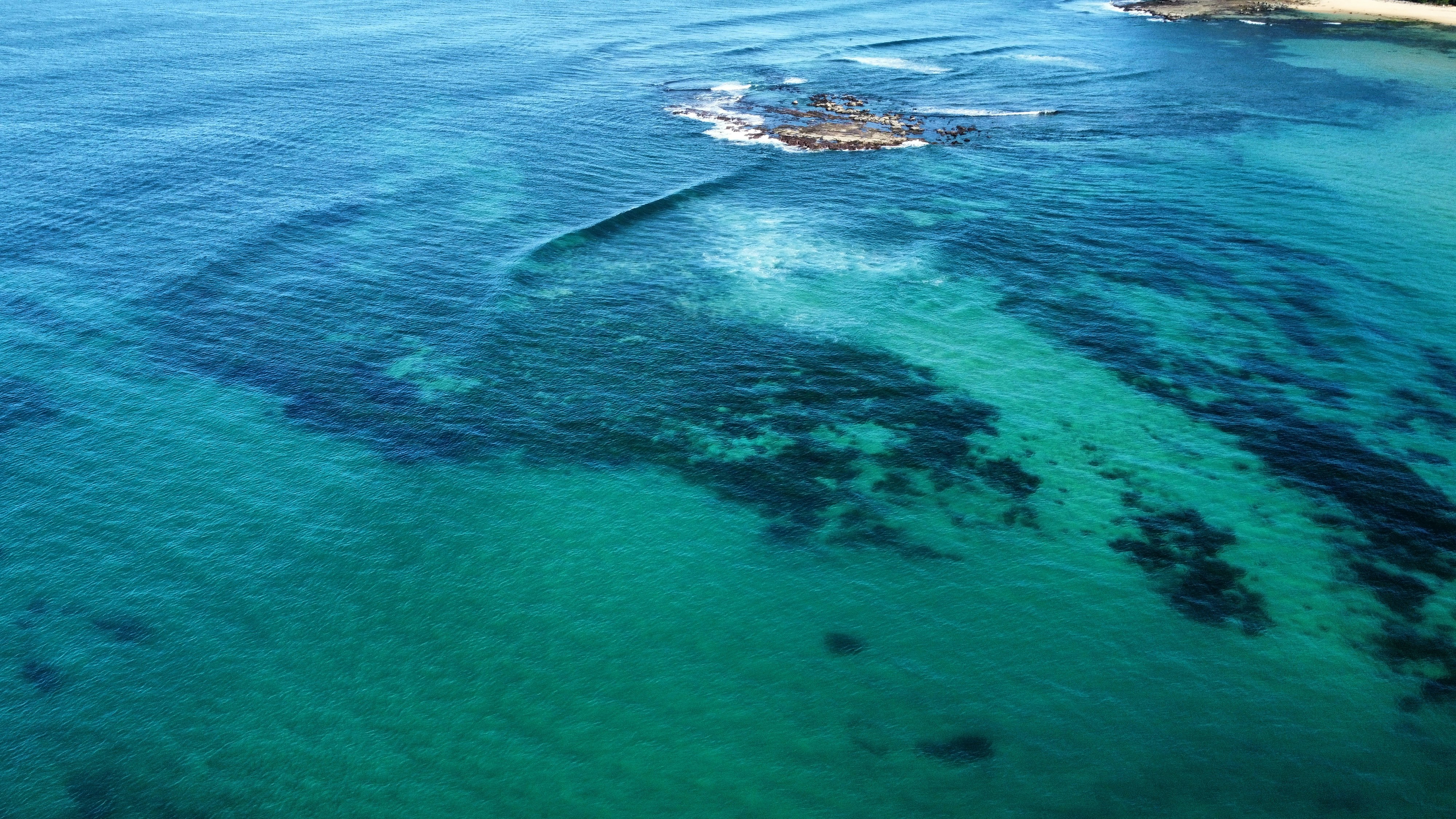 Azure ocean waters reveal turquoise shallows and dark coral patches, captured from above to showcase the seabed contours and vibrant color variations. The shot emphasizes a broad seascape with gentle gradients and subtle wave texture.