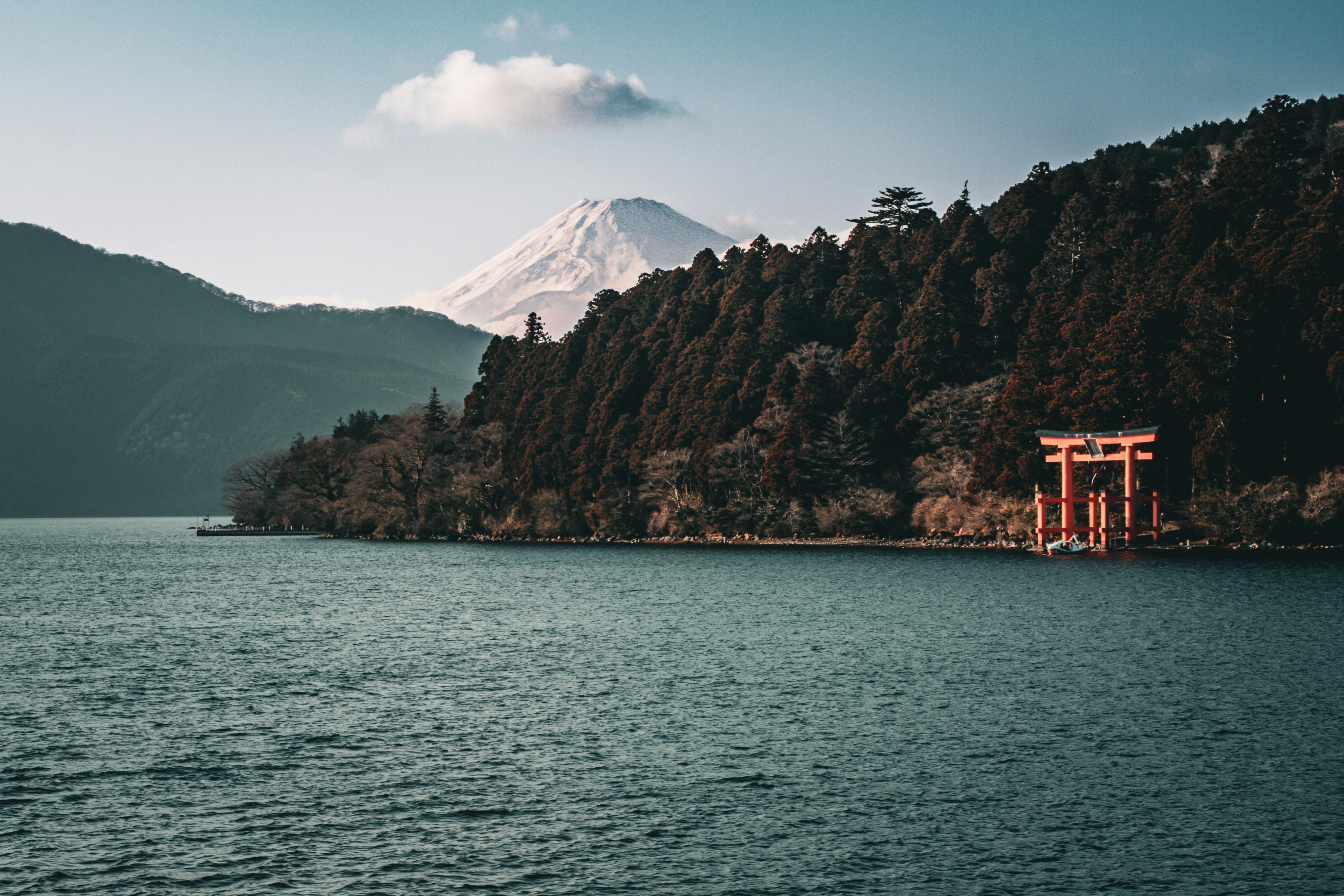 Coastal lake with a distant snow-capped mountain and a bright red torii gate along a tree-lined shore; the scene emphasizes tranquil landscape and cultural landmark.