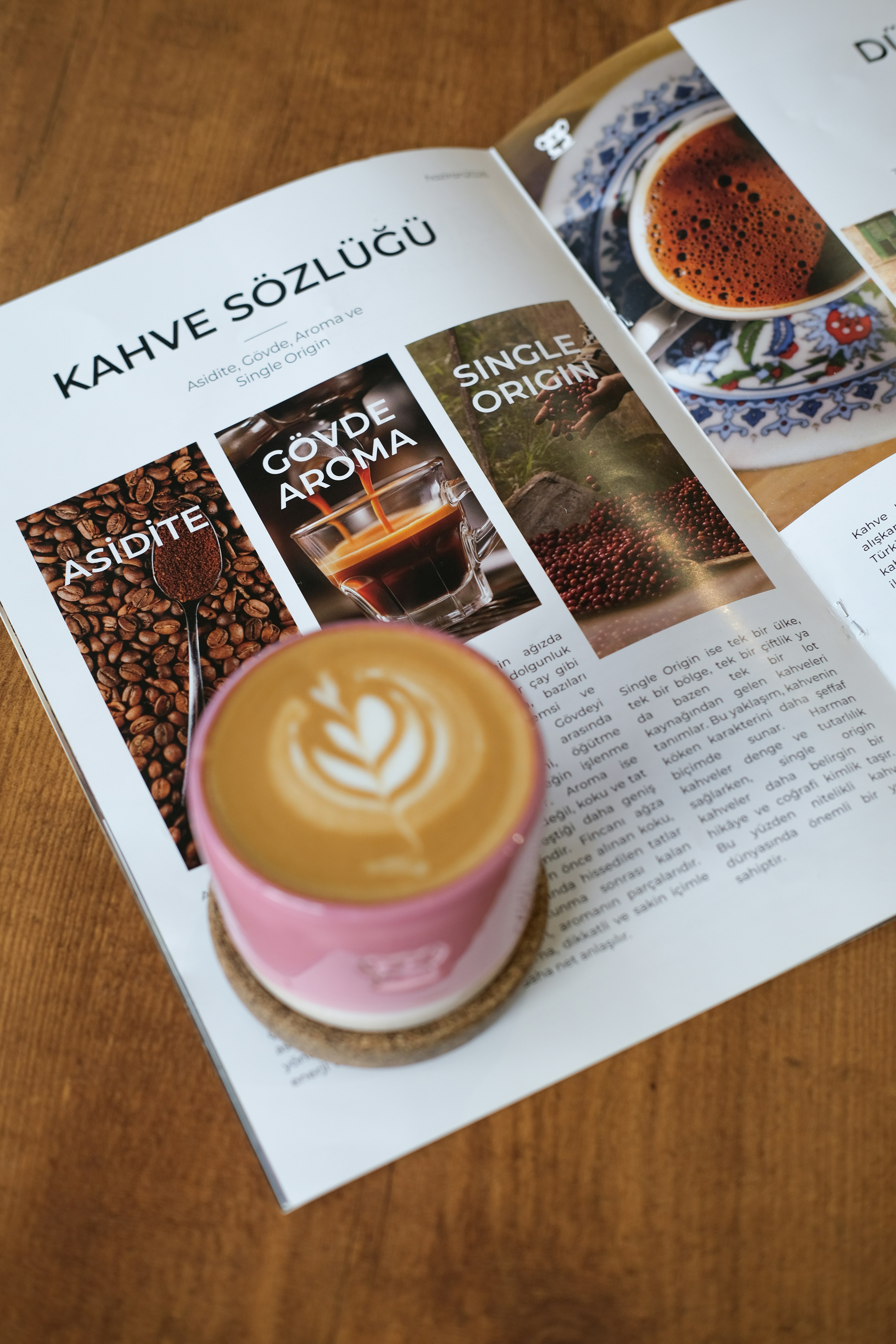 A glossy magazine page featuring coffee-themed panels rests on a wooden table, with a pink cup of latte perched atop a coaster in the foreground.