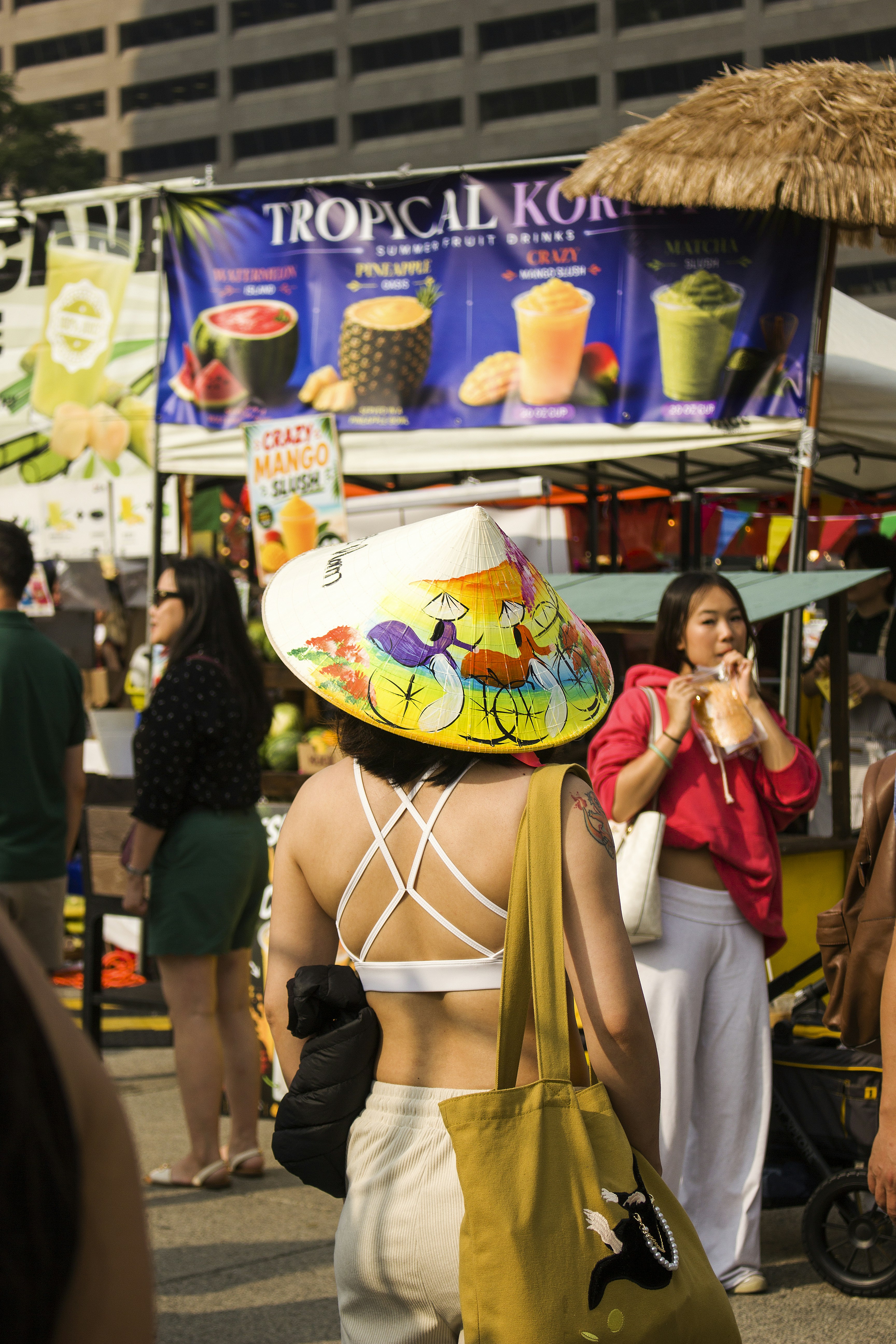 Vibrant street festival scene with a woman wearing a decorative conical hat, white crisscross top, and a yellow bag amid colorful market banners and shoppers.