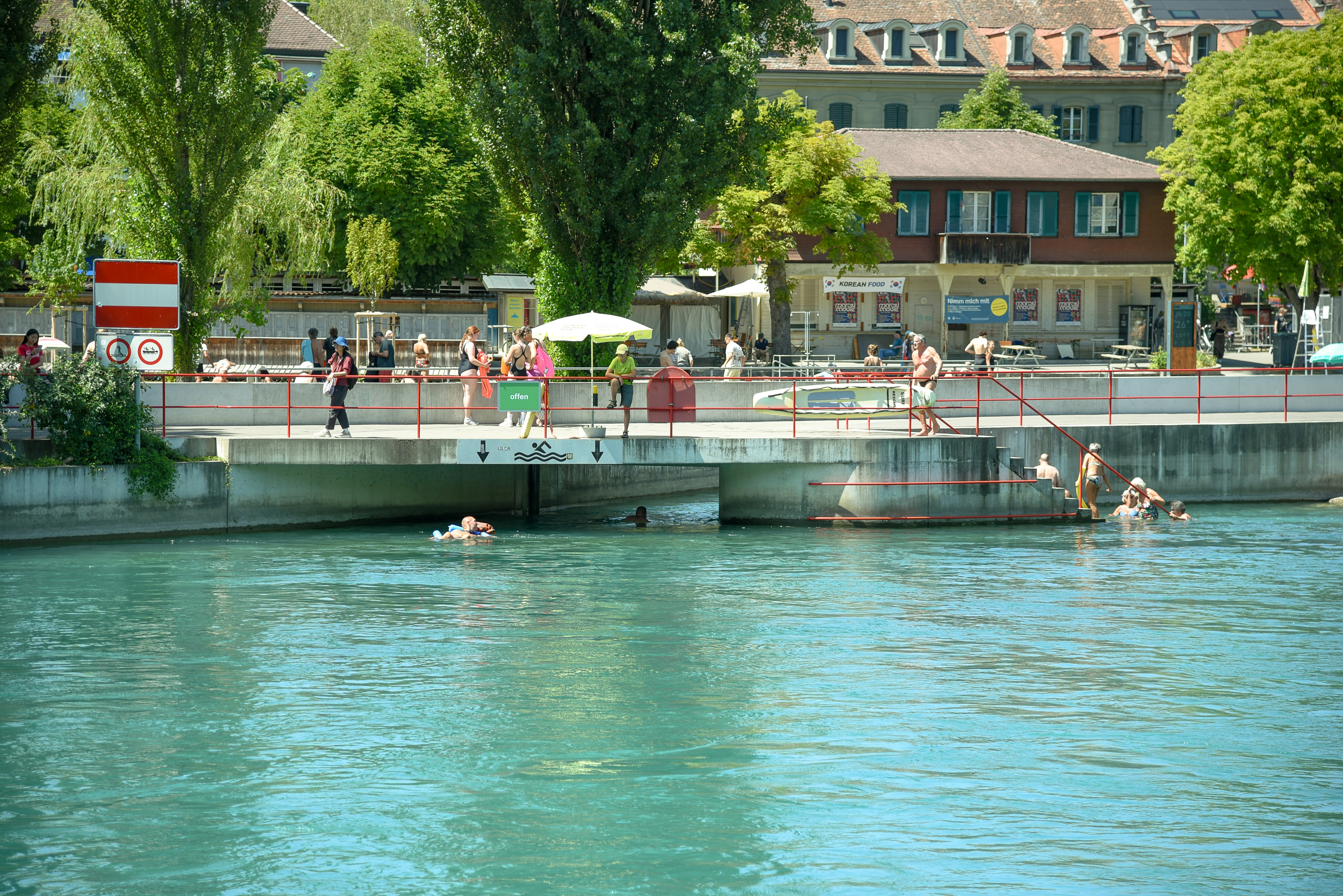 People by the turquoise riverbank under a shaded promenade