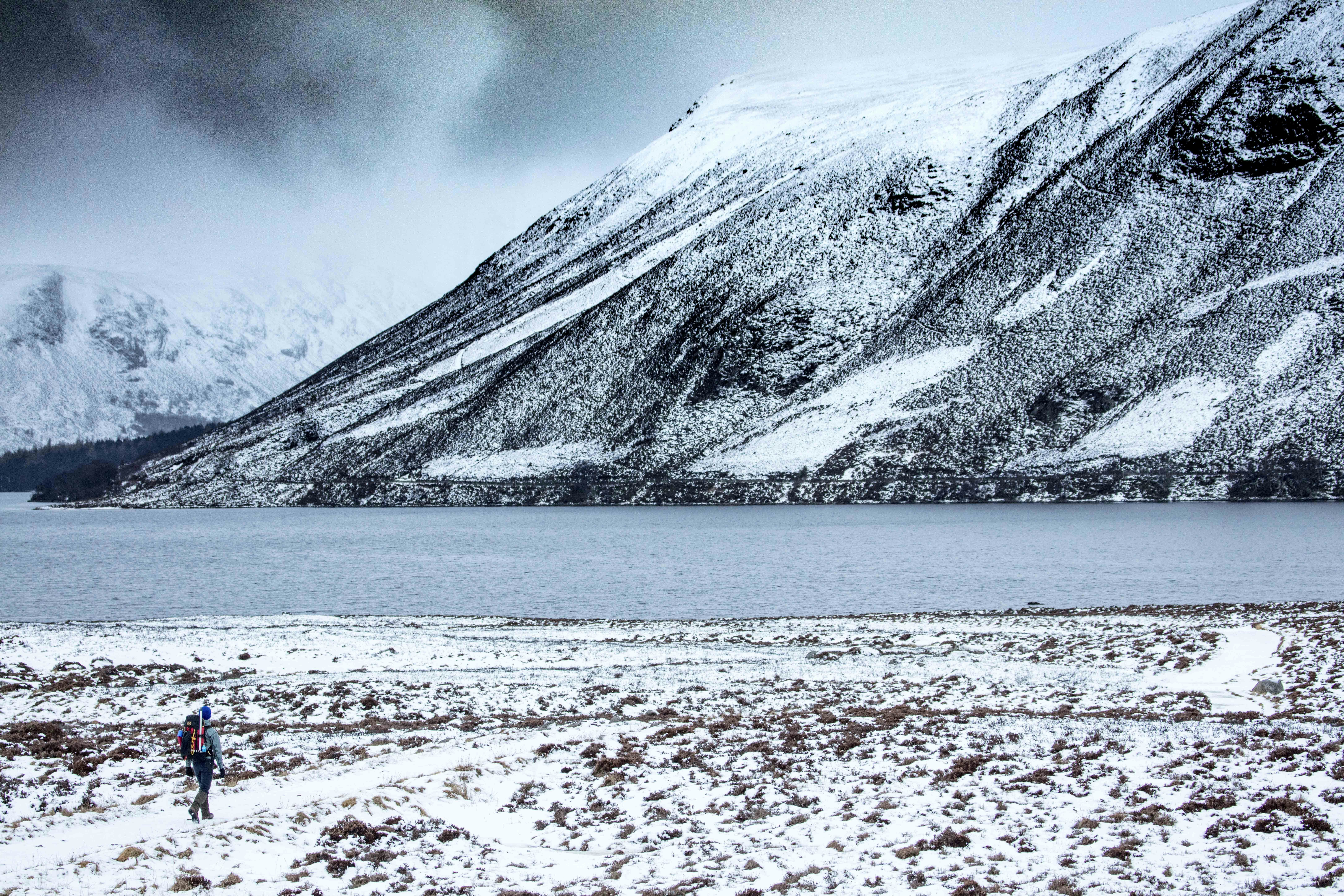 Snowy shoreline with a lone hiker near the water's edge, framed by vast, rugged mountains and a cloudy sky; a landscape photograph capturing scale and cold atmosphere.
