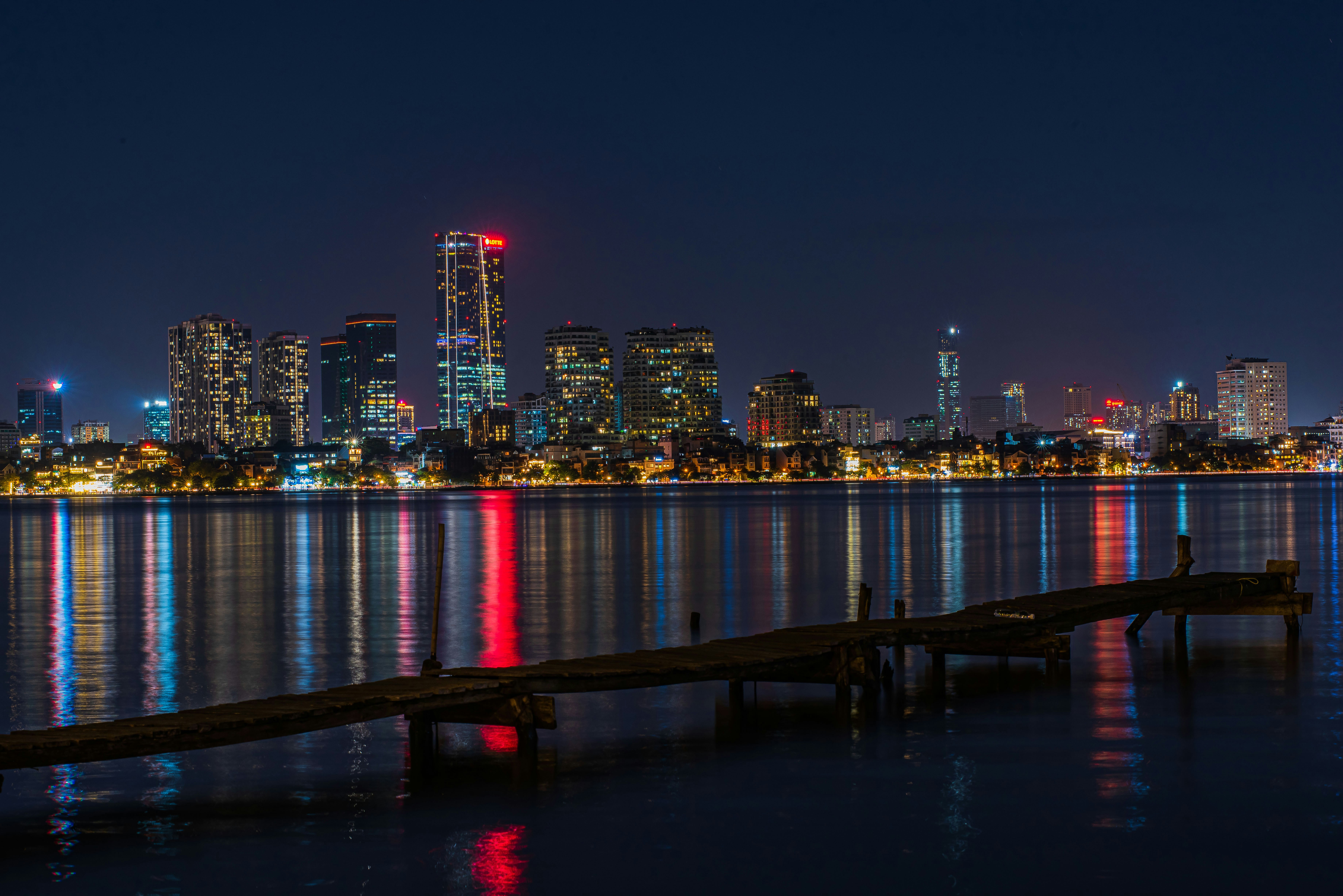 Nighttime city skyline glows across a calm harbor with reflections; a weathered pier stretches into the water, anchoring the foreground.