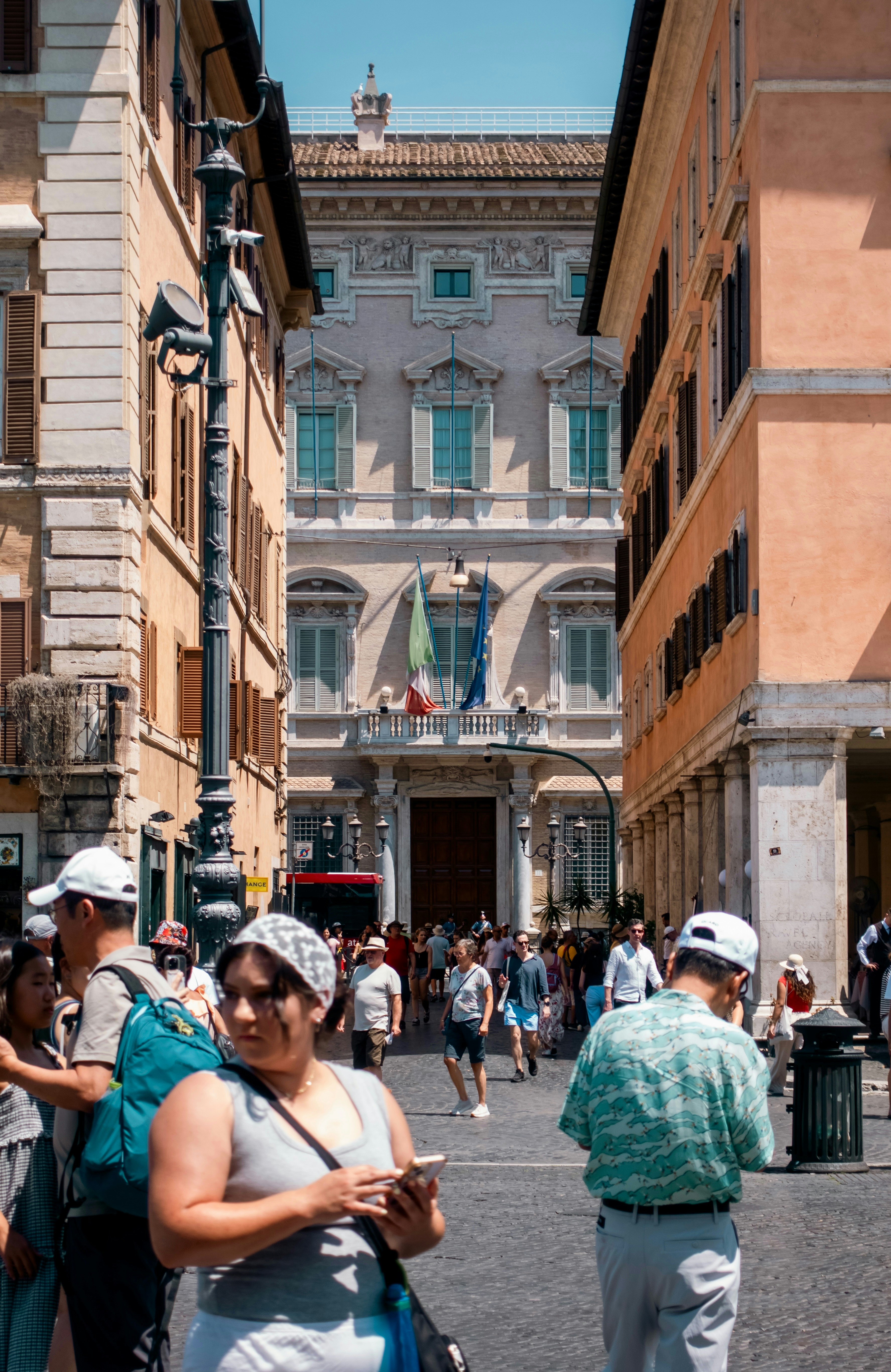 Crowded cobblestone street framed by warm-toned historic buildings; pedestrians pause and stroll under a clear blue sky, with a grand doorway and flags visible in the background.