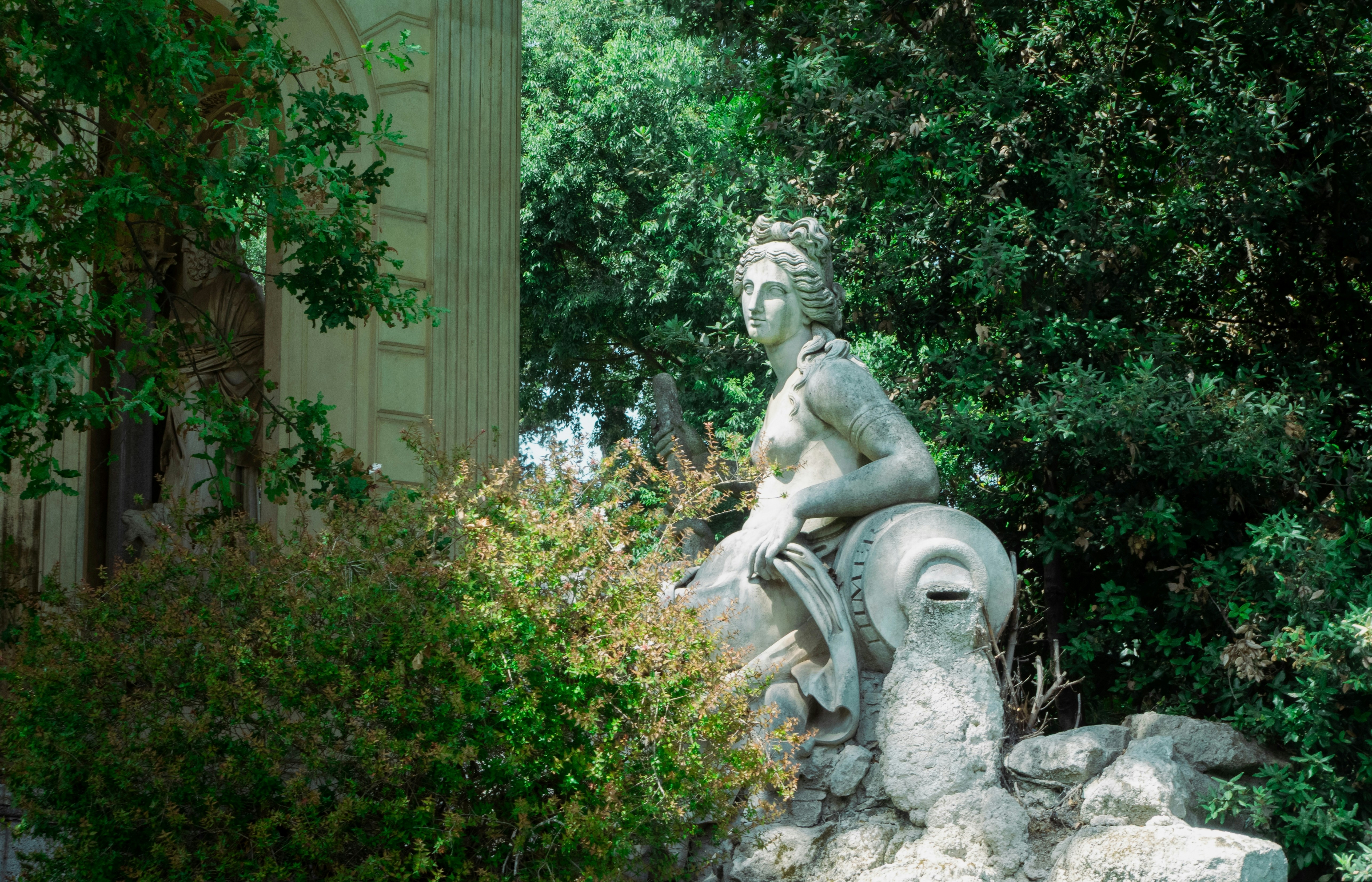Stone garden statue bathed in dappled sunlight among lush shrubbery and trees, with classical helmeted figure perched atop weathered scrollwork. The scene conveys serene, courtyard ambience.