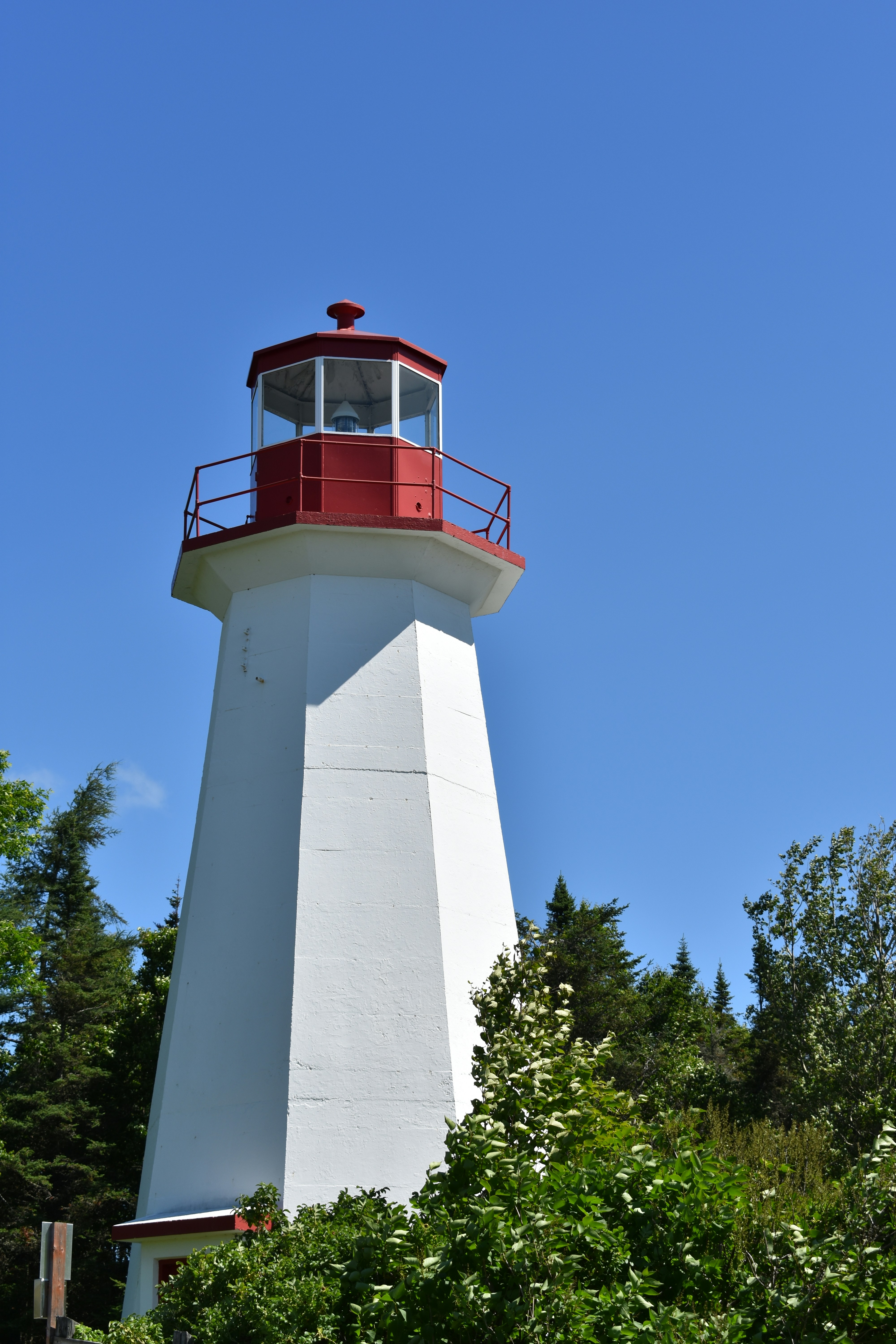 White lighthouse with red lantern and railing stands tall against a clear blue sky, surrounded by green foliage.