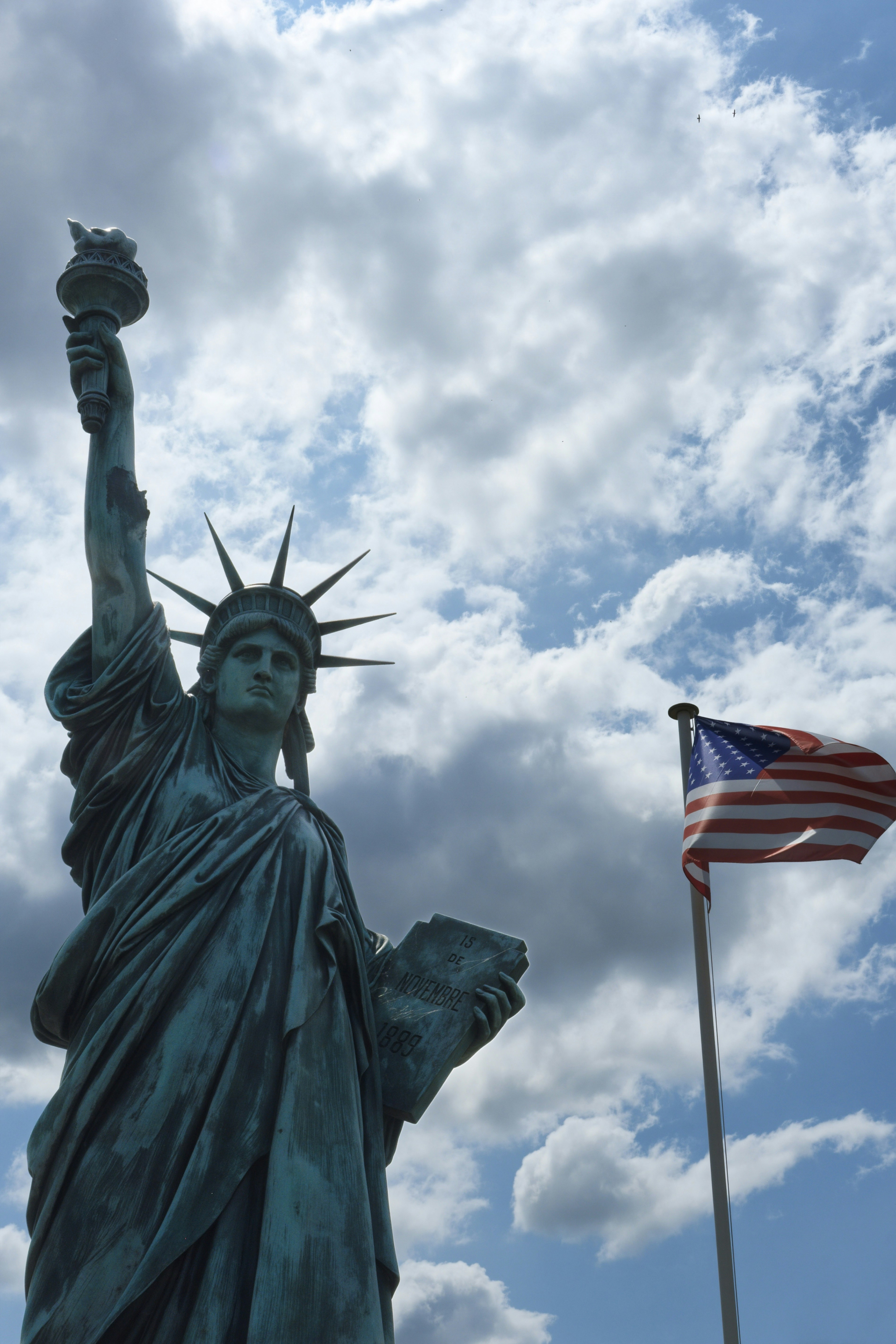 Statue of Liberty stands tall with American flag waving nearby under cloudy sky - The Statue of Liberty is seen holding a torch high while an American flag flies close by. The scene captures clouds in the sky, adding depth to the view of this landmark.