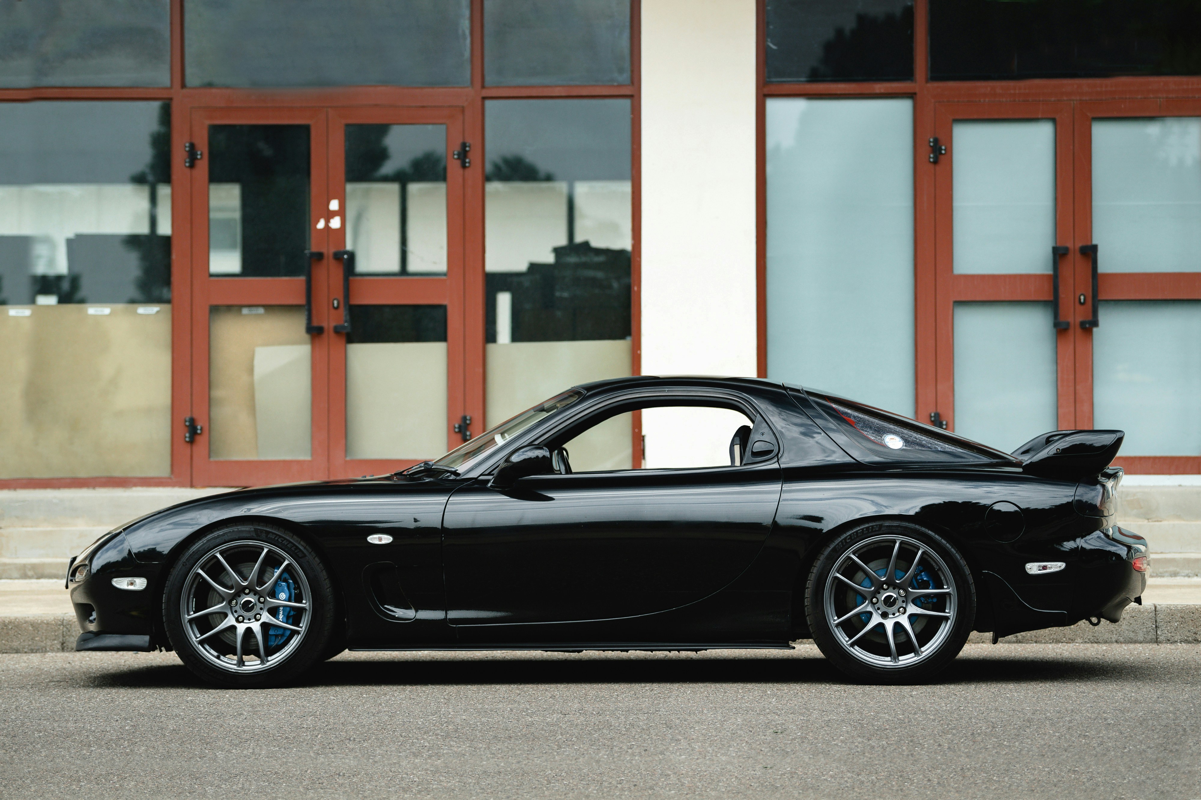 Glossy black sports coupe parked along a curb with a modern building backdrop; emphasis on wheel detail and sculpted body lines.