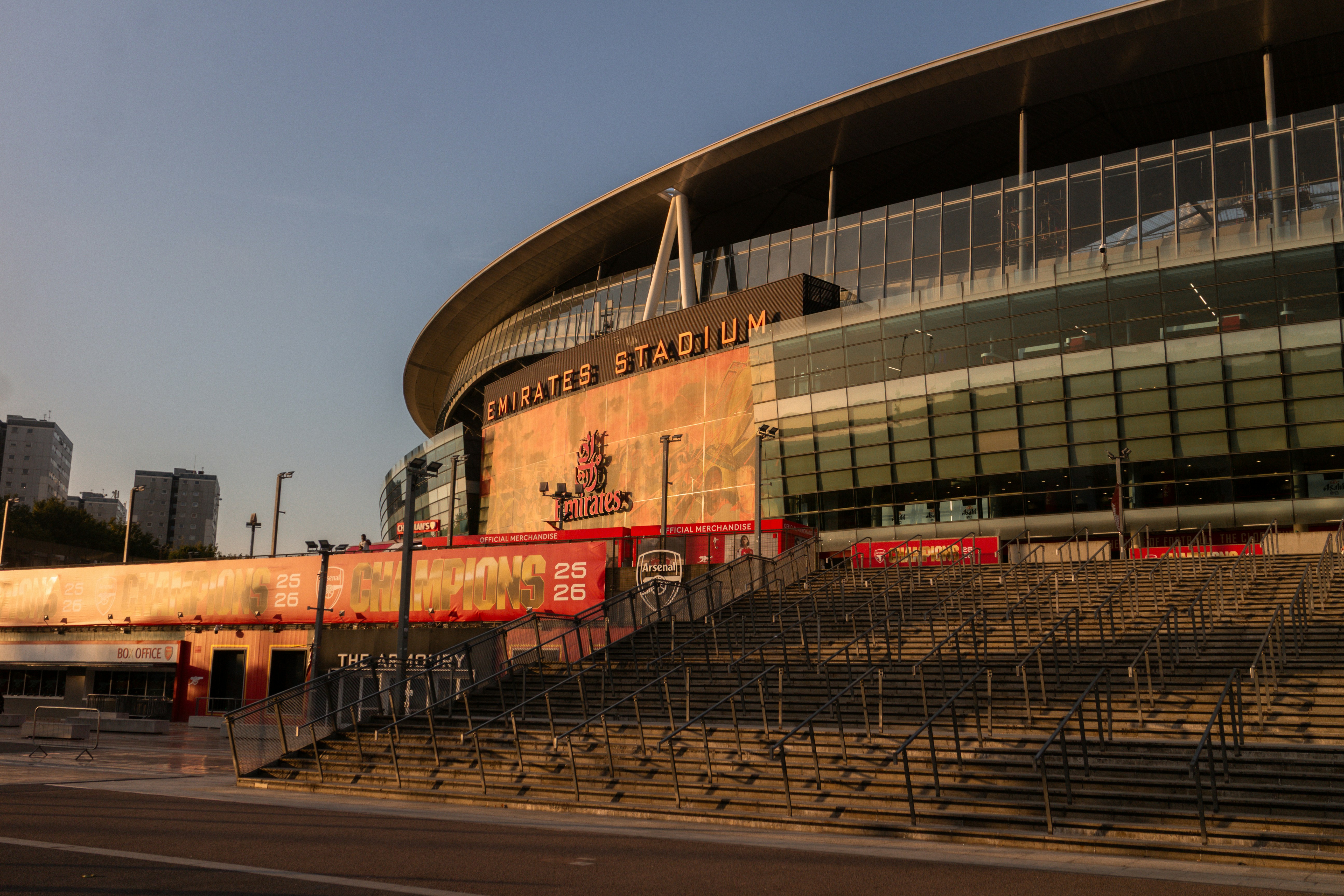 Wide-angle view of a modern stadium facade with glass and concrete steps, bathed in warm late-afternoon light. The scene emphasizes the architectural curve and empty bleachers.