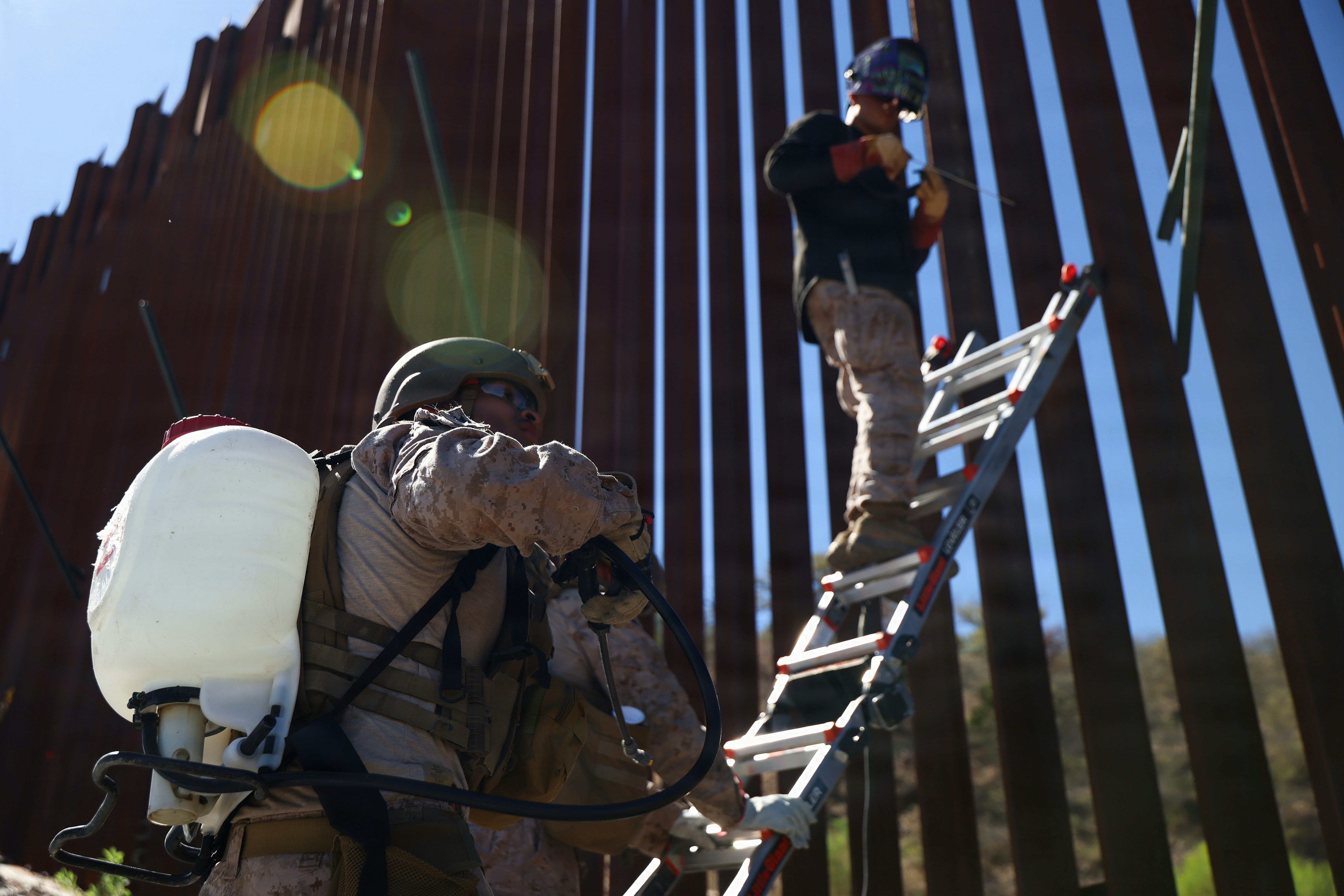 Two soldiers in protective gear work on a steel barrier, one on a ladder and another at the base with a backpack sprayer, against vertical slats under bright daylight.
