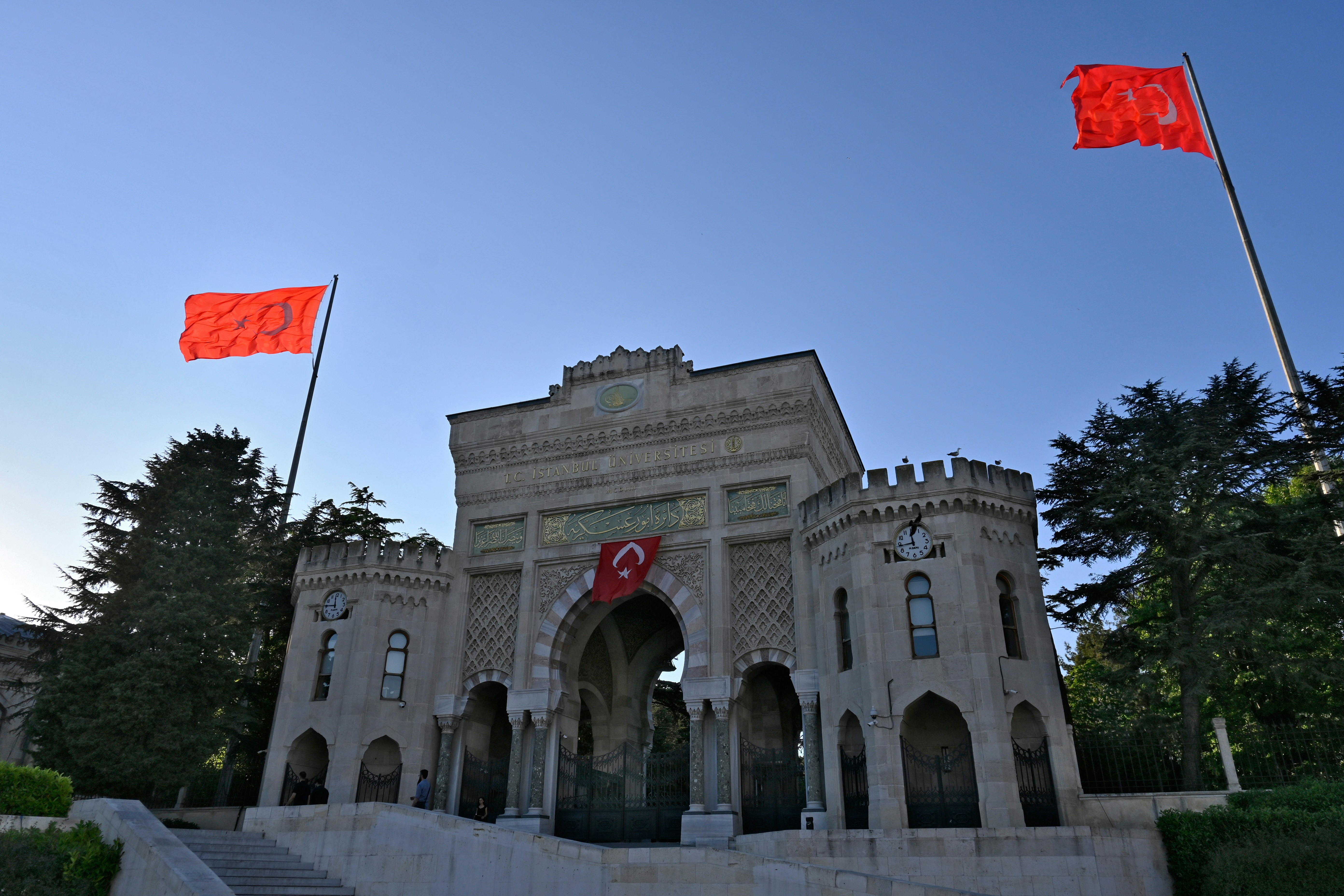 Stone gate with arched opening and decorative reliefs, flanked by two flags and trees; a grand architectural entrance set on steps. Logo- or emblem-like stonework prevalent, center framed by open sky.