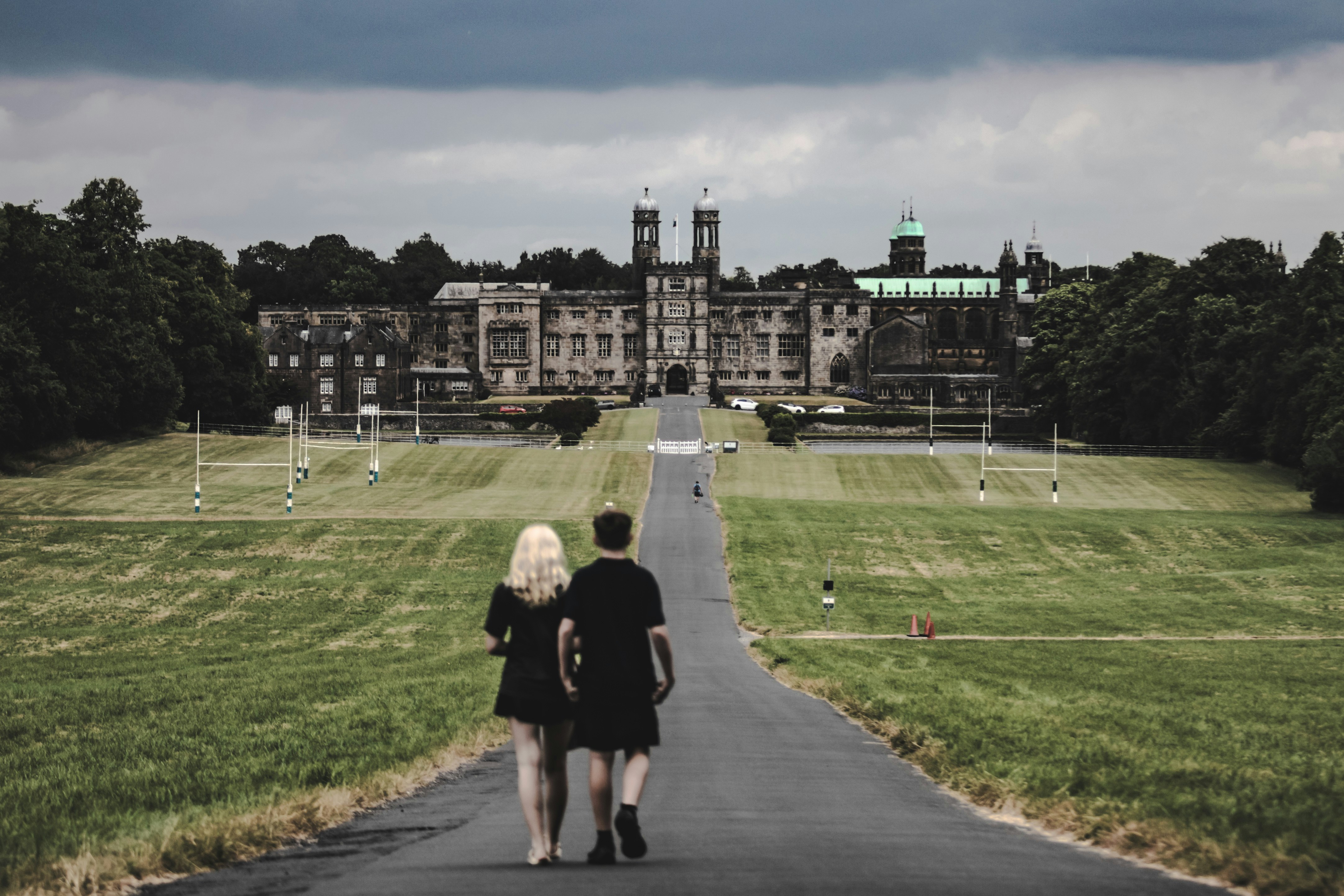 A couple walks along a central path toward a grand, distant palace on a wide, manicured lawn. The scene emphasizes symmetry and depth as the figures recede toward the historic structure.