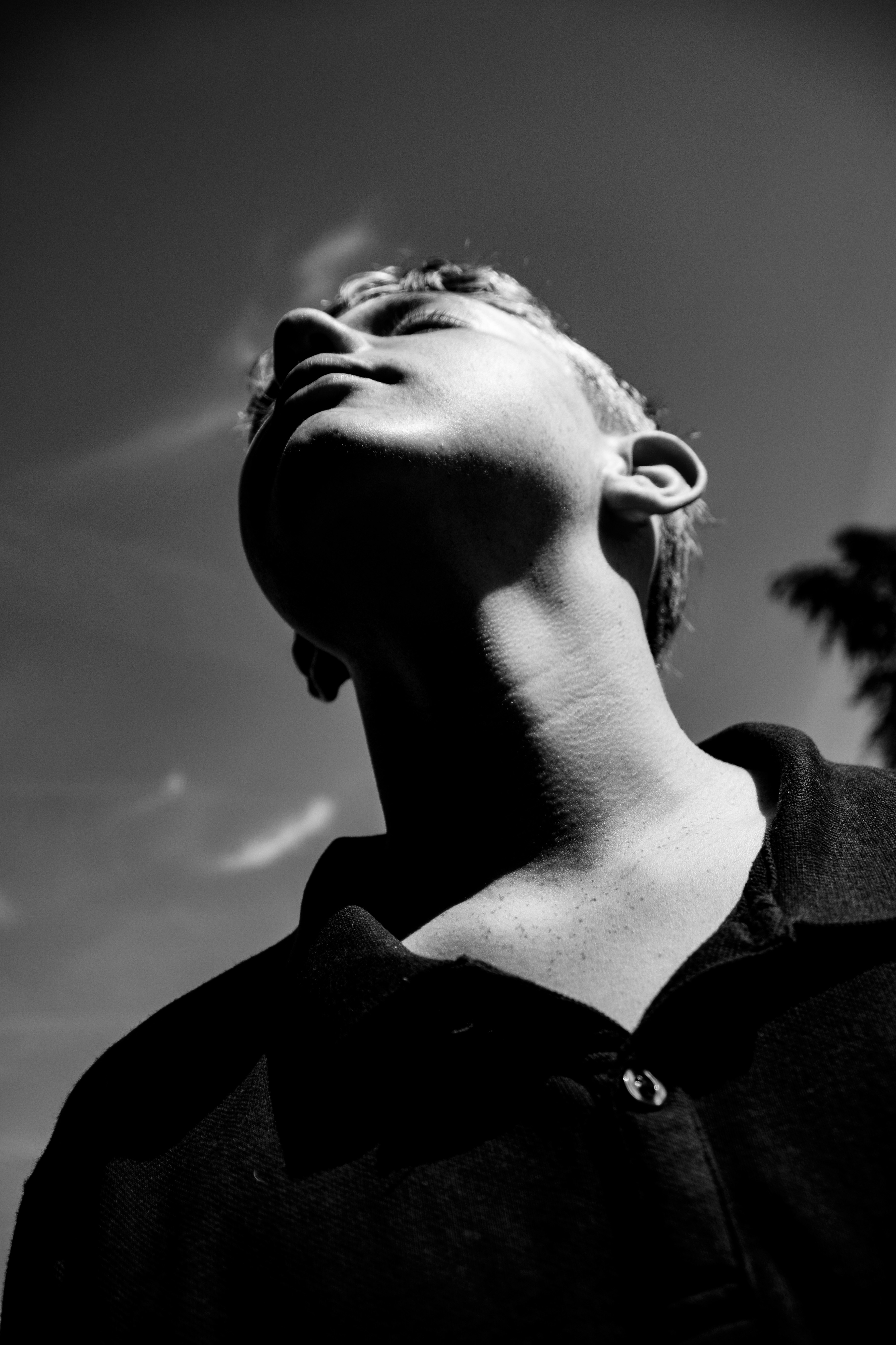High-contrast black-and-white portrait of a young man angled upward against a cloudy sky; minimalist composition focusing on the jawline and neck. Logo-free, photographed as a moody, documentary-style head-and-shoulders capture.