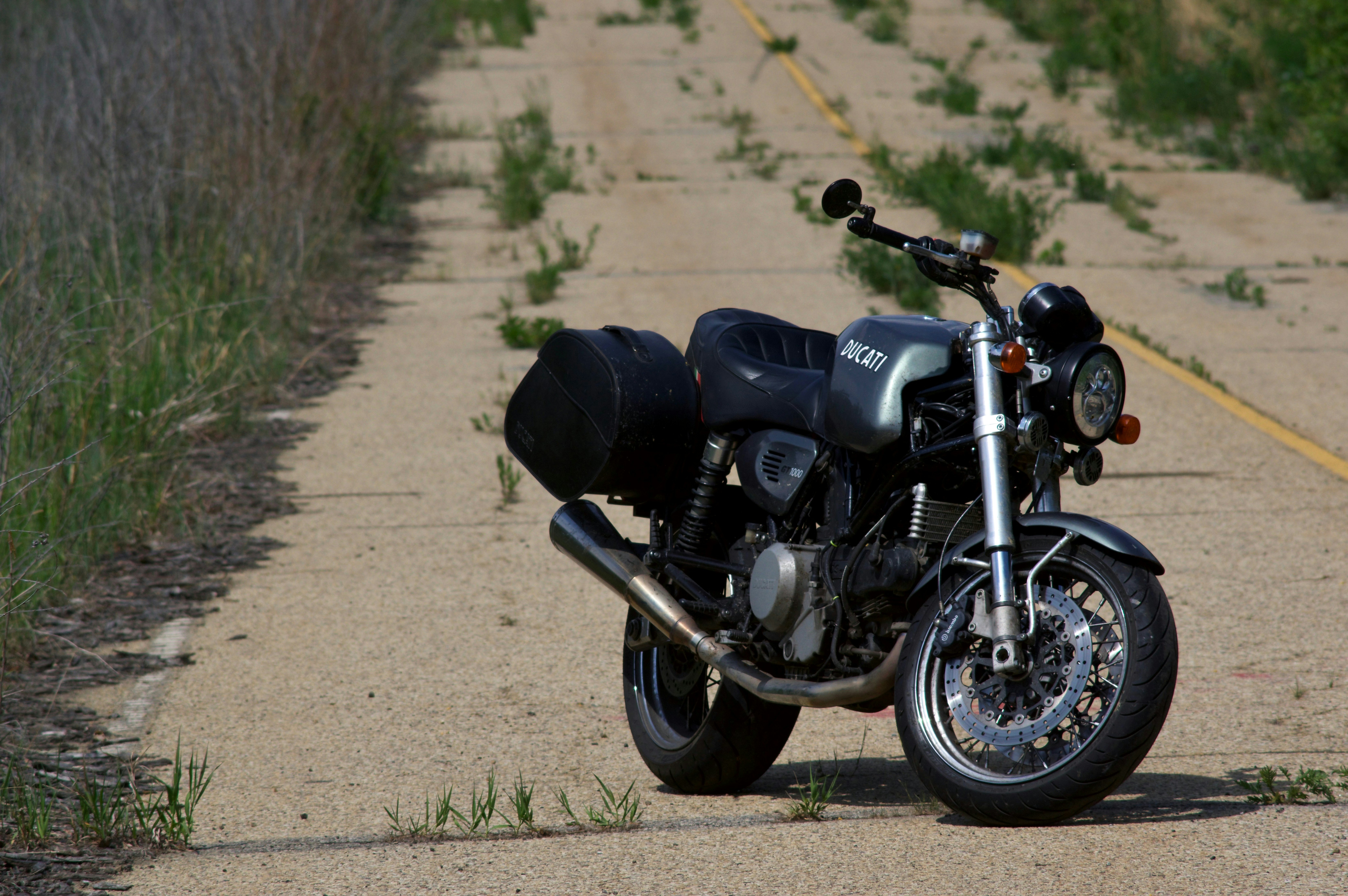 A vintage Ducati motorcycle sits on a cracked pavement with overgrown weeds along the edges, highlighting a rugged, retro aesthetic. The scene emphasizes the bike’s silhouette, metal finish, and the desolate, industrial setting.