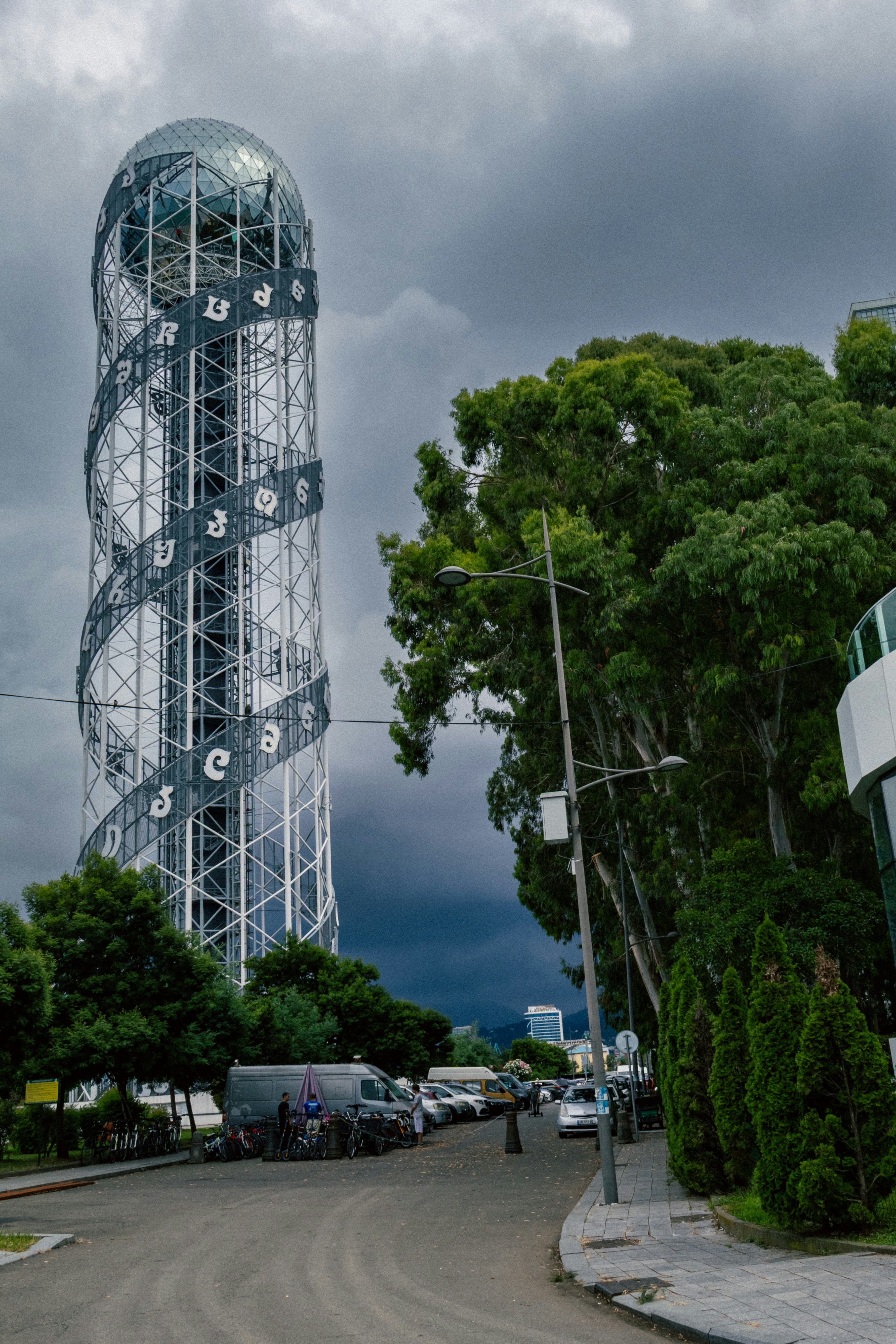 Skeletal tower rises beside a tree-lined street with parked vehicles and a paved walkway, set against a moody, overcast sky. A logo-like spiraling structure dominates the left, framing a quiet urban scene.