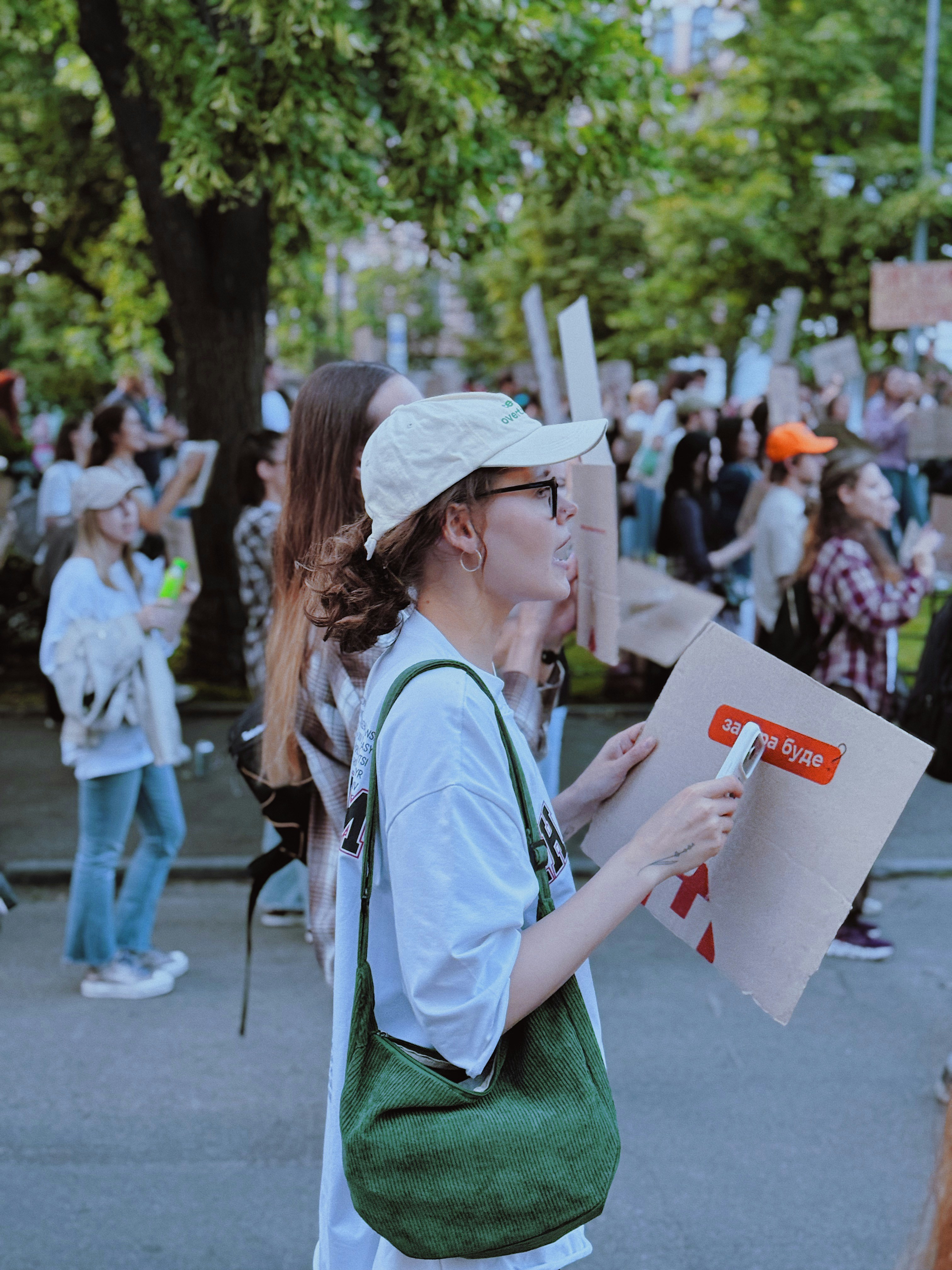 A young woman wearing a white cap and glasses holds a cardboard sign at a crowded outdoor event; she carries a green bag and wears a light blue shirt, with other participants and trees in the background.