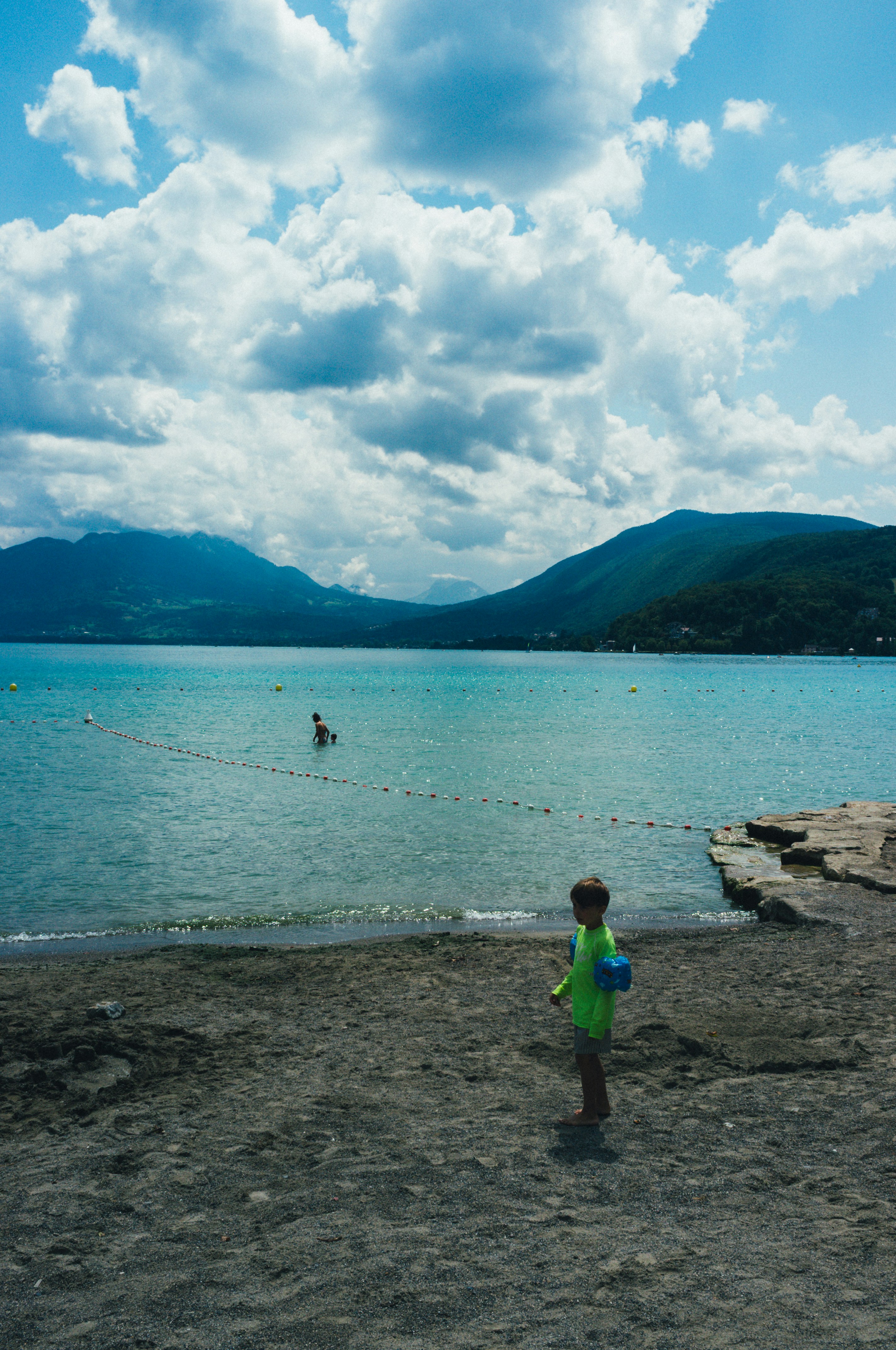 Child in bright green shirt stands on rocky shore looking toward the water; a swimmer is visible in the distance as mountains meet a blue lake. A calm, outdoor scene without a formal logo or artwork.
