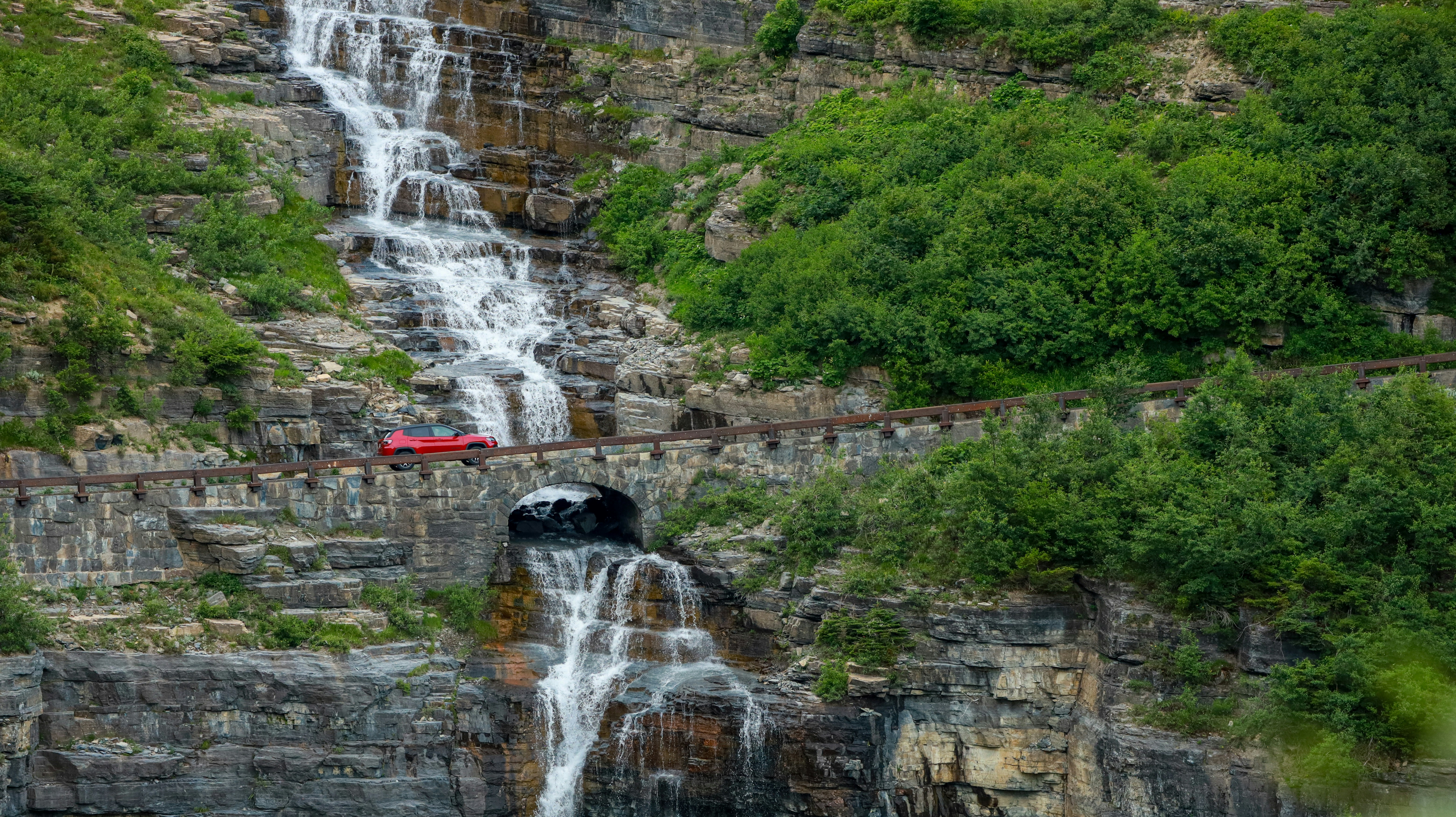Cascade beneath the stone viaduct: a red car on a cliffside bridge
