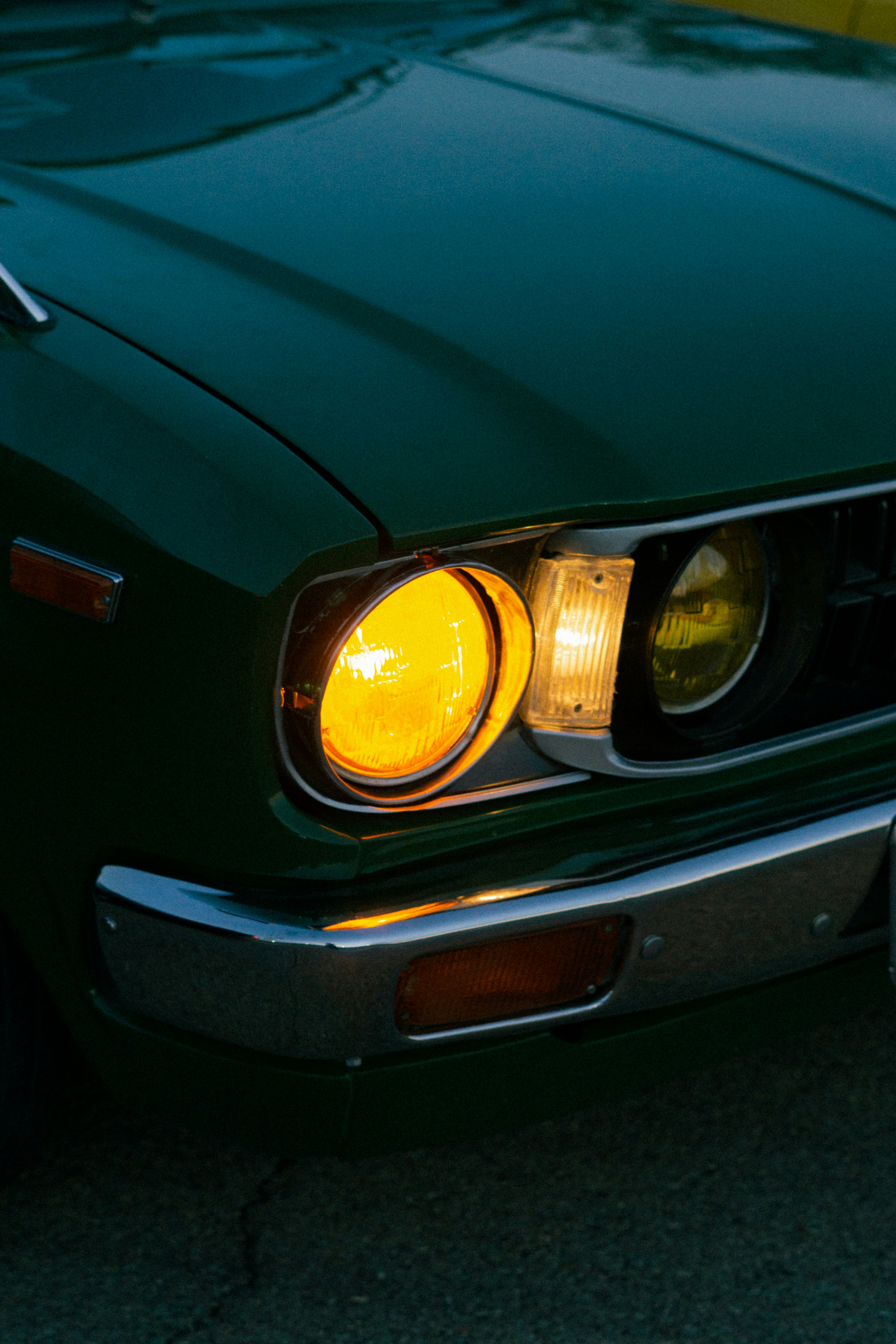 Close-up of the car's front end showing the illuminated amber headlight and surrounding grille; the scene emphasizes vintage chrome and rich green paint. This is a photographic capture of a real vehicle exterior.