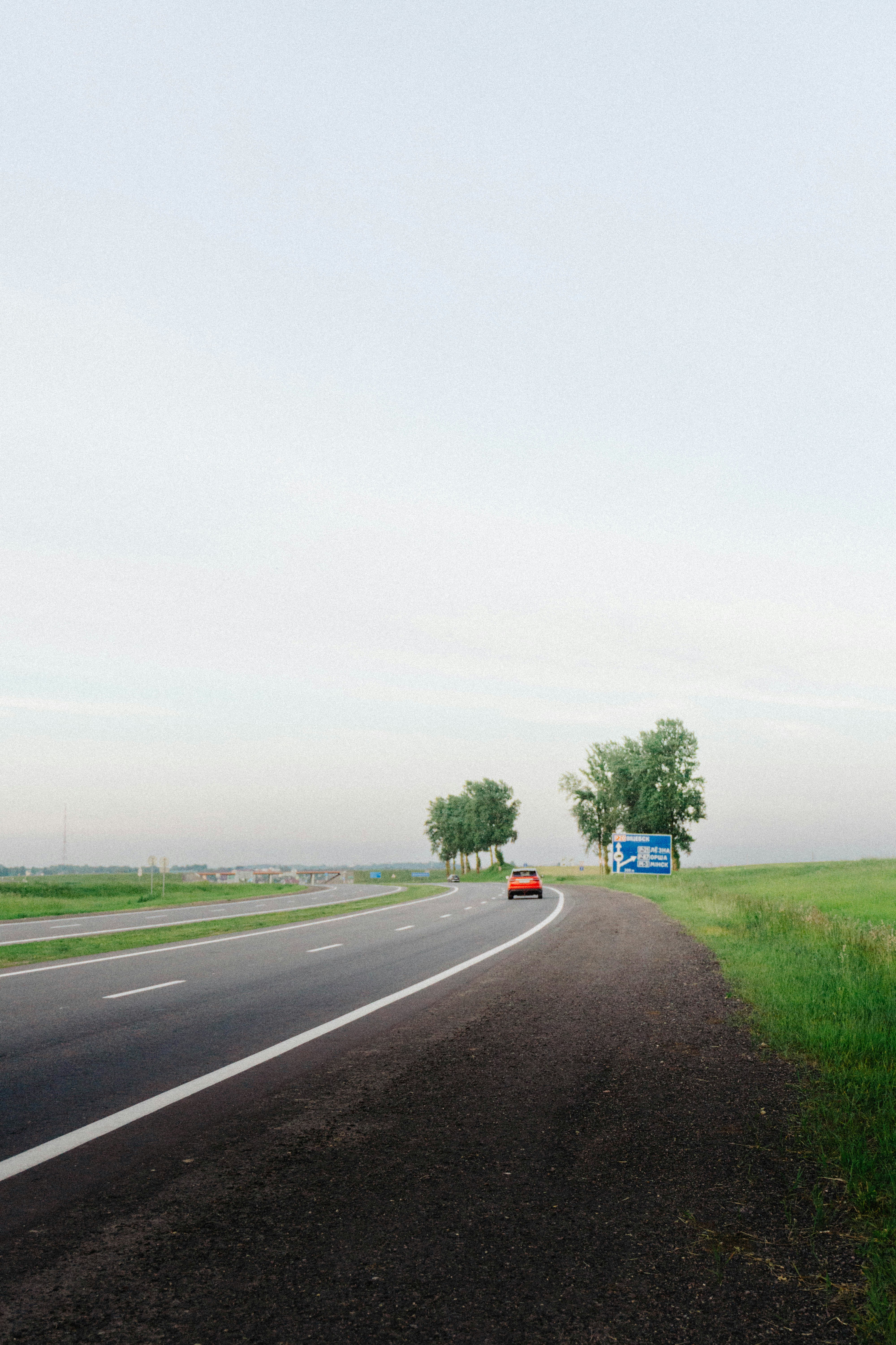 A red car travels along a two-lane highway that curves toward a grassy verge and distant trees under a pale blue sky. The scene emphasizes open countryside and road perspective.