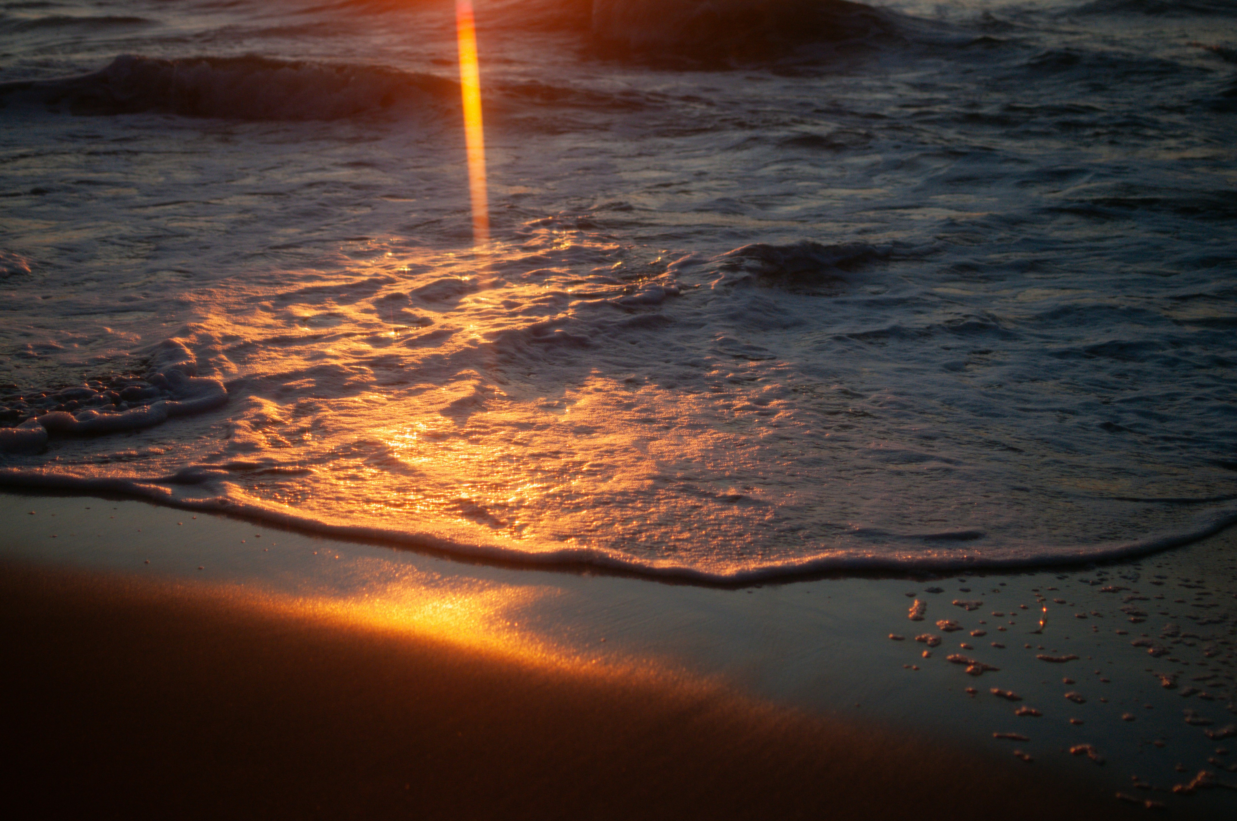 Warm glow along the shoreline with reflected sunset light on gentle waves and wet sand. A calm coastal scene highlighting a low-angle sunbeam and shimmering water texture.