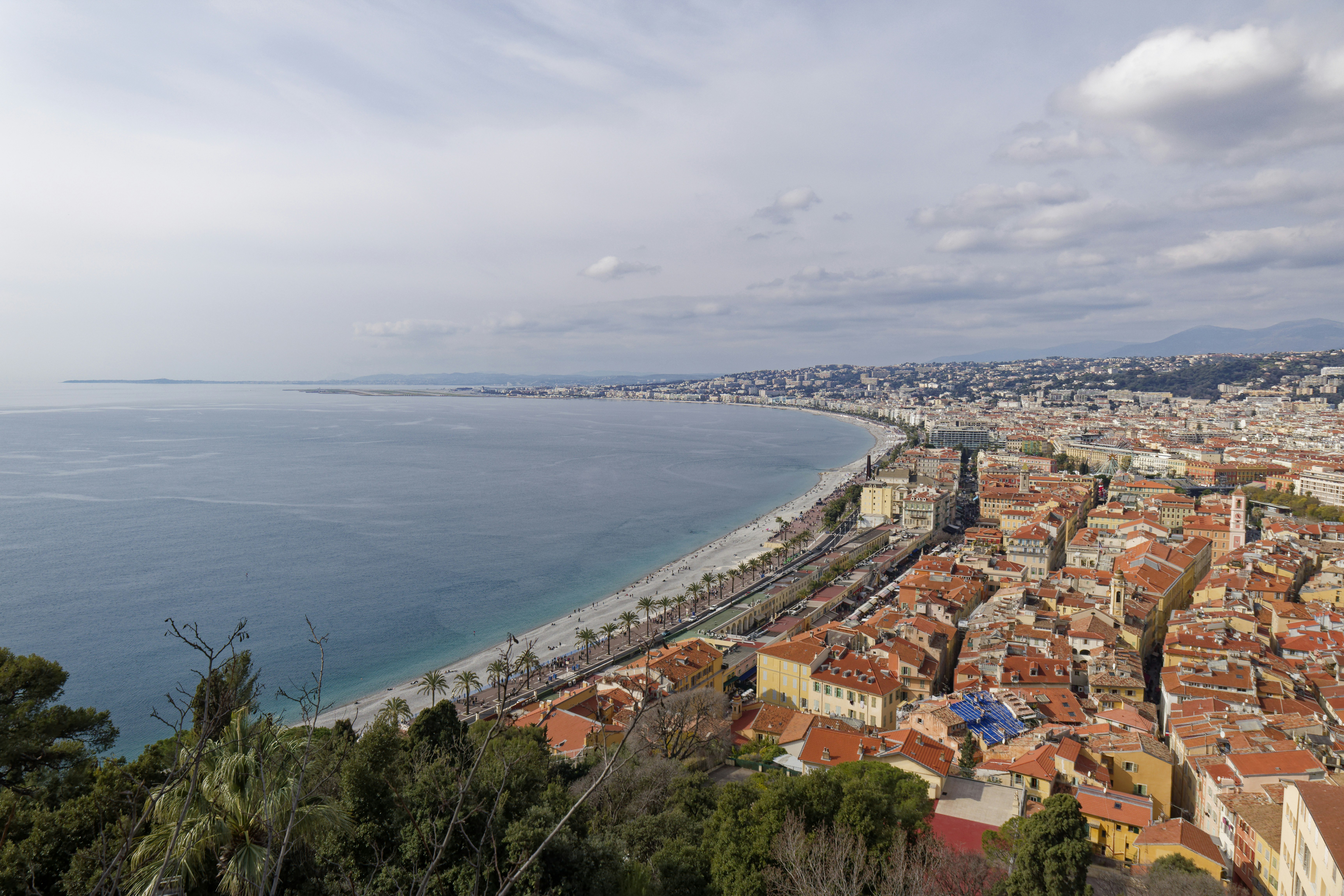Coastal town hugs a curved shoreline with terracotta roofs cascading down a hillside toward the sea. A promenade lines the water while distant hills frame the horizon.